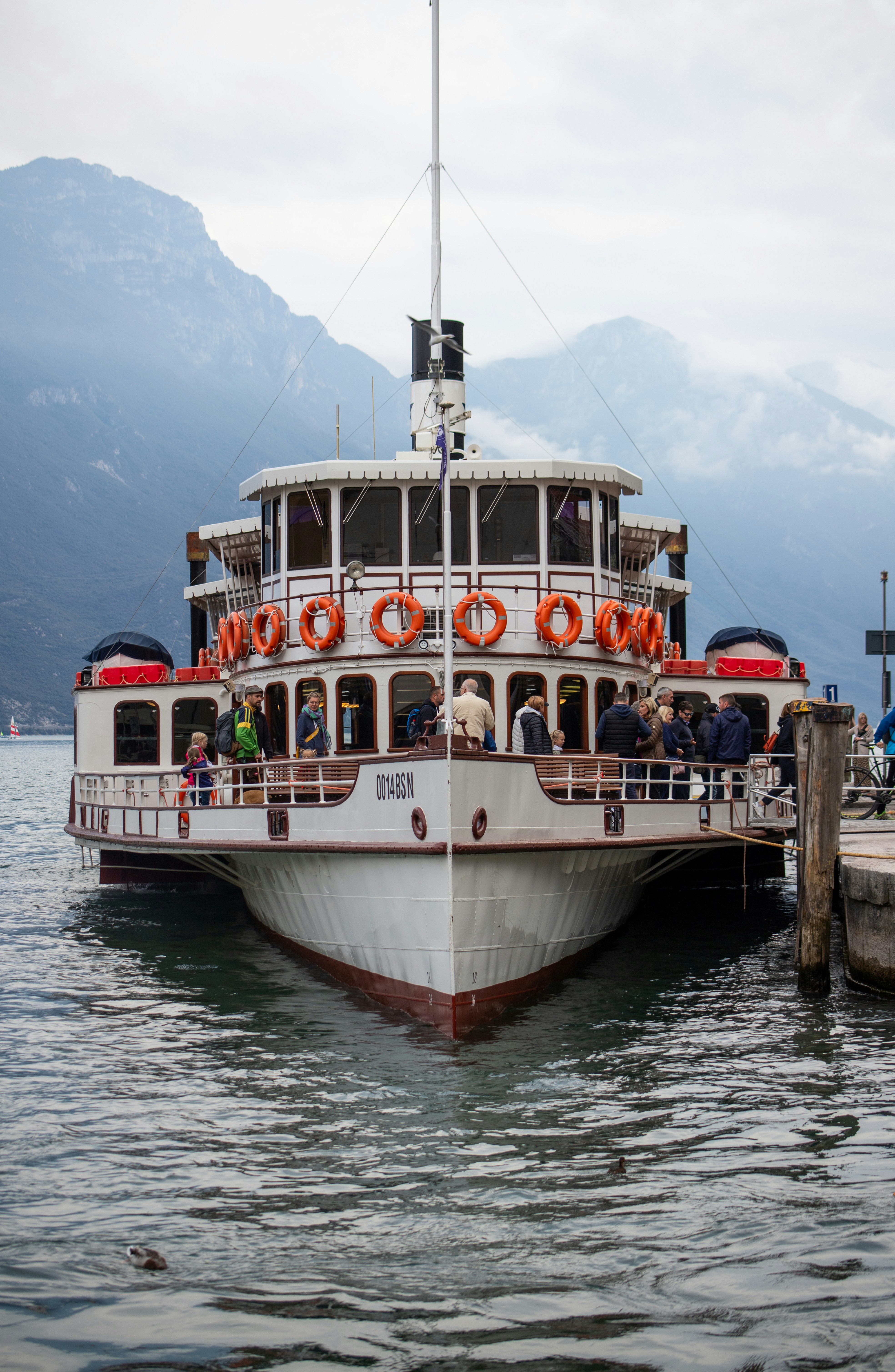 Historic boat docked at a serene lakeside, surrounded by towering mountains and passengers boarding. 