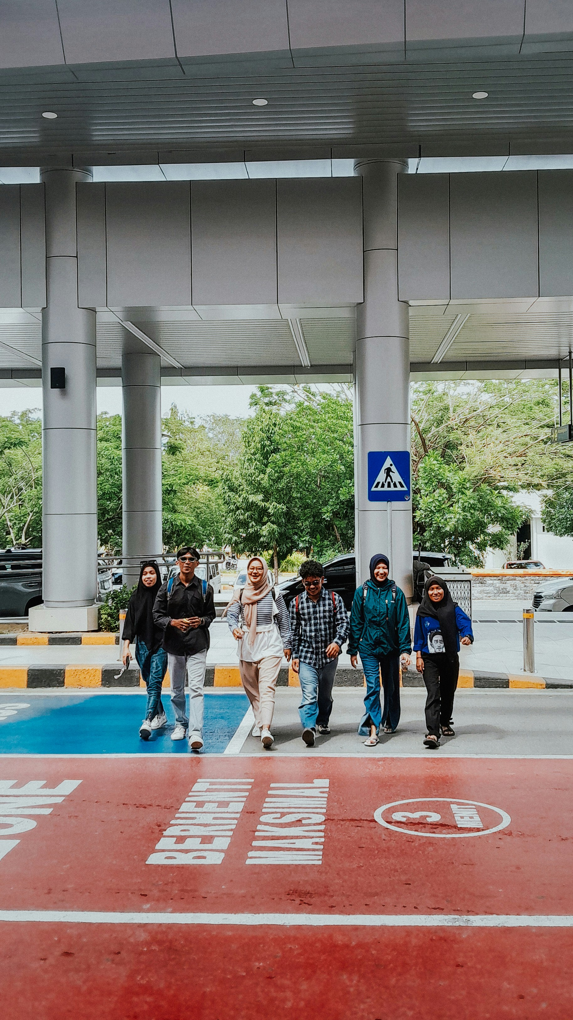Seven friends walk across a painted crosswalk under a modern transit canopy, framed by tall columns and a pedestrian sign.