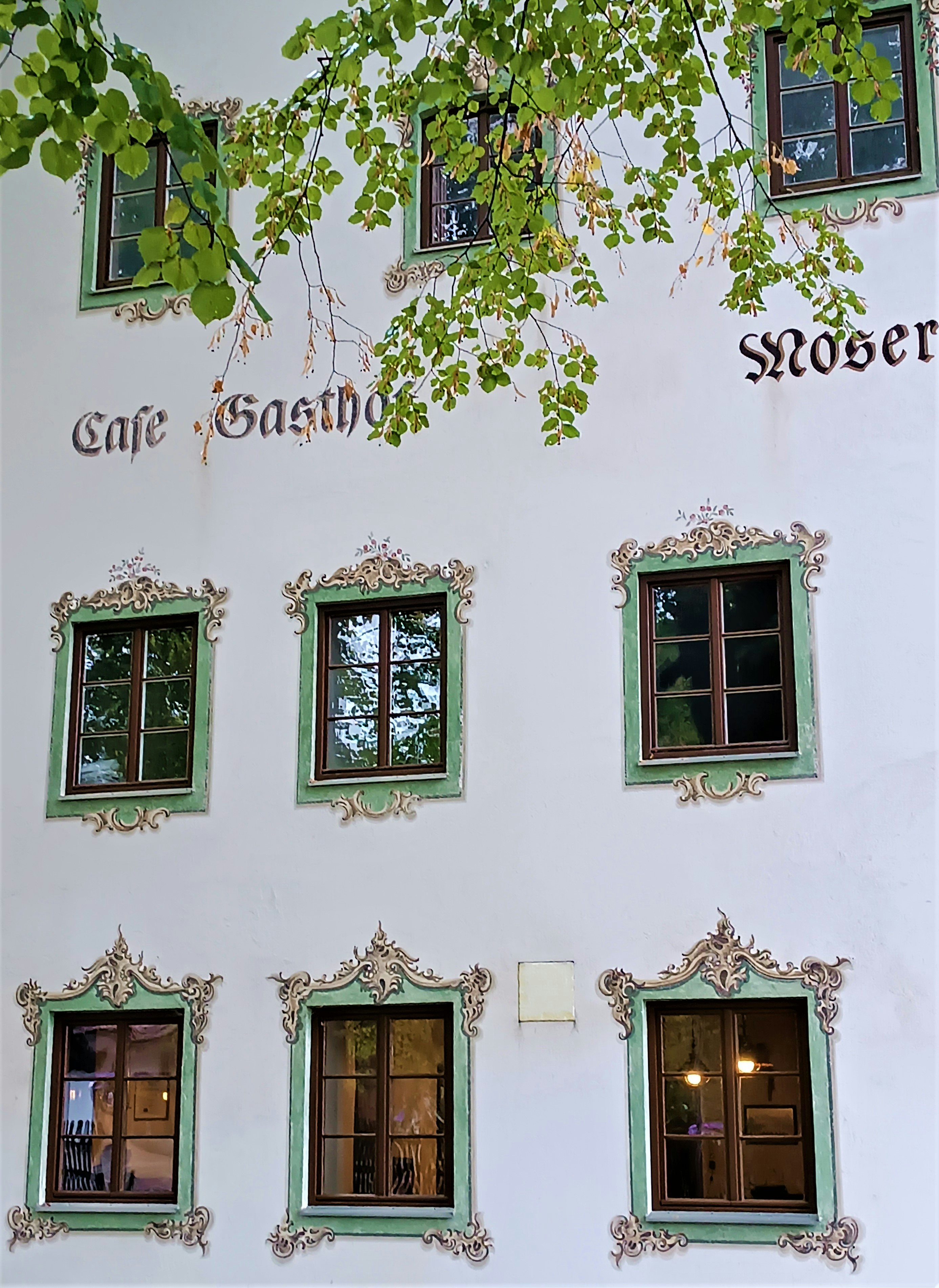 Photograph of a white façade with ornate green-trimmed windows and hanging vines. The scene emphasizes architectural detail and a quaint café vibe.