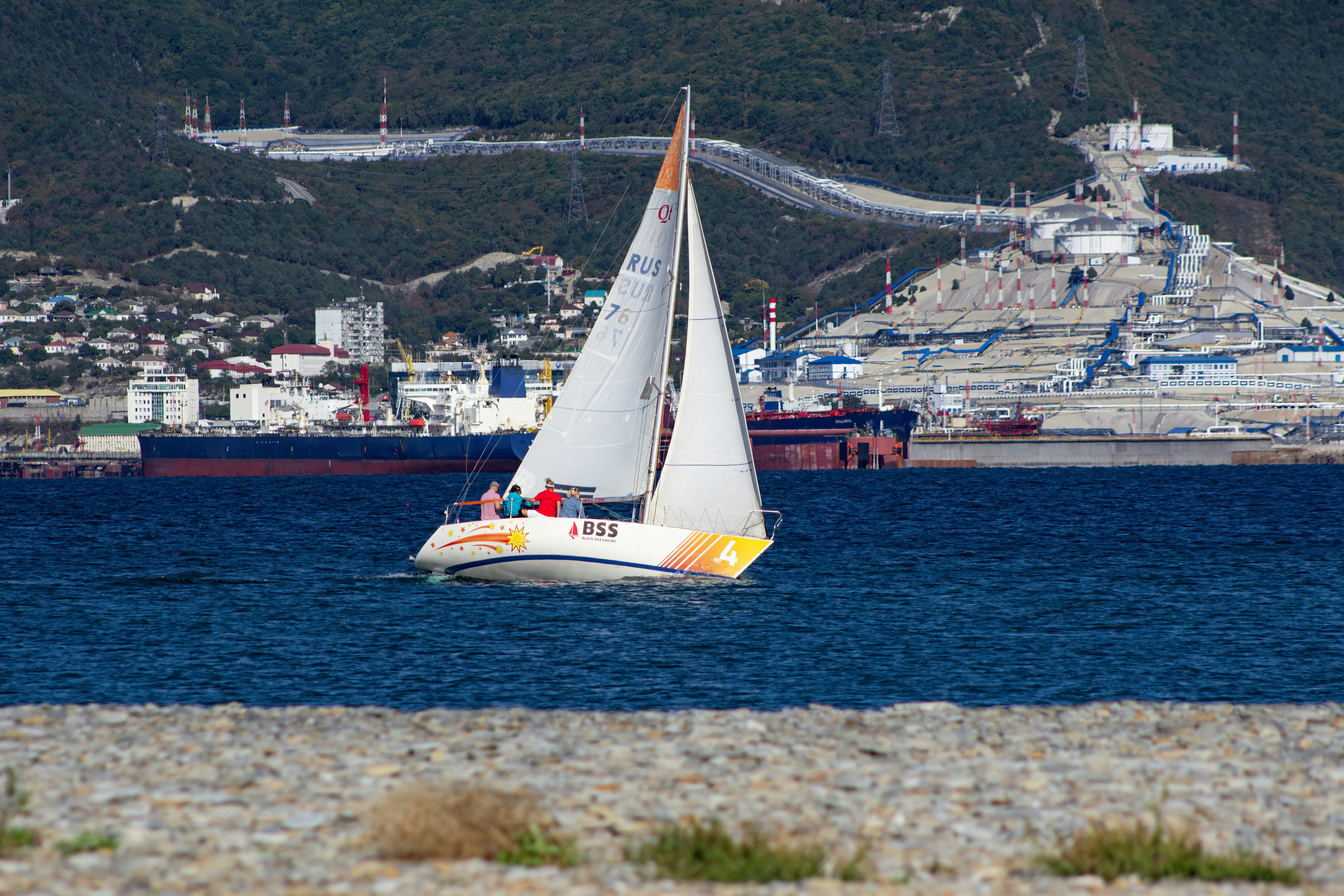 White Sails, Steel Horizons: A Sailboat Gliding Past a Port City and Hills