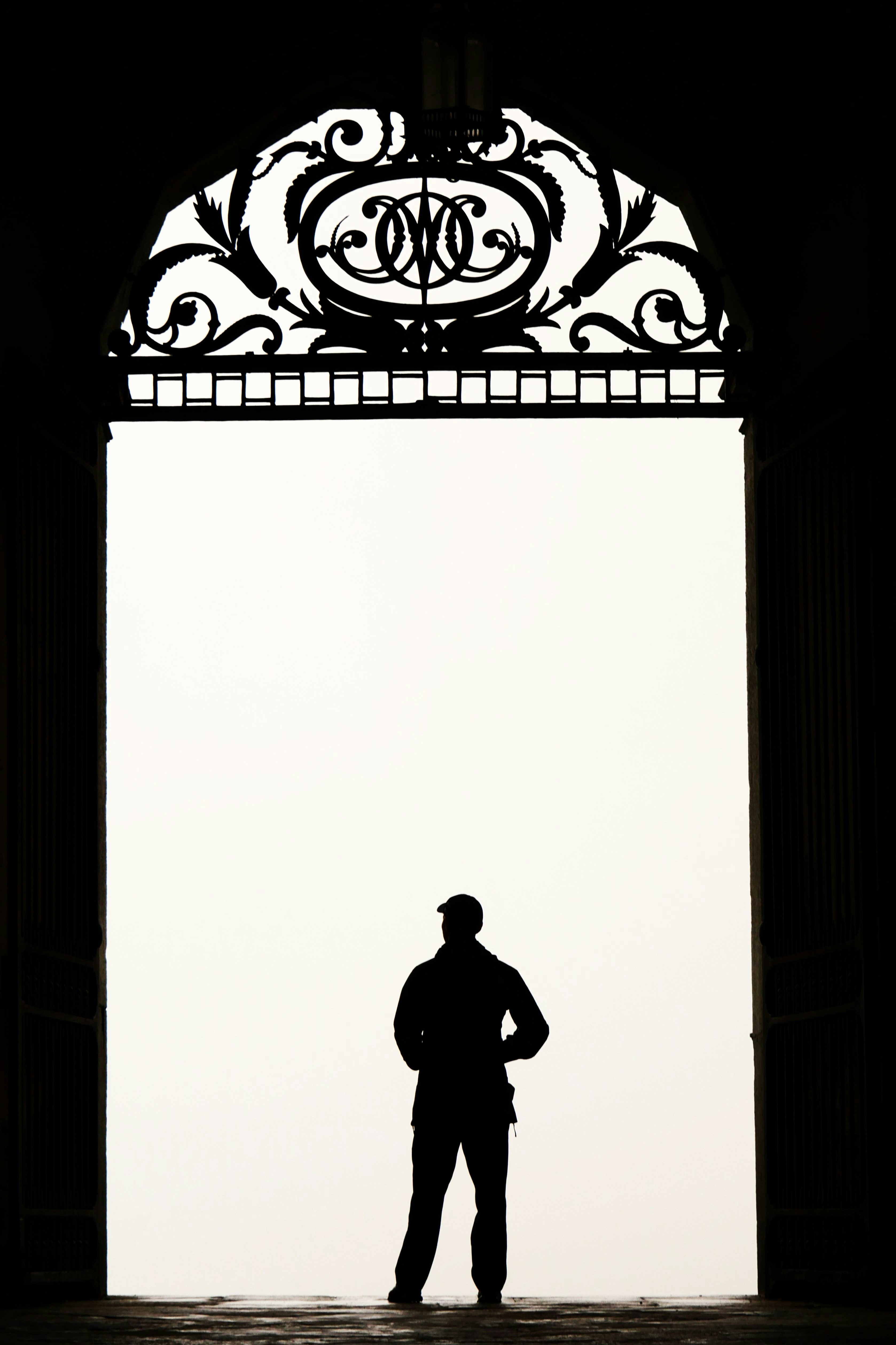Silhouette of a lone figure framed by a large, backlit doorway with an intricate wrought-iron arch.