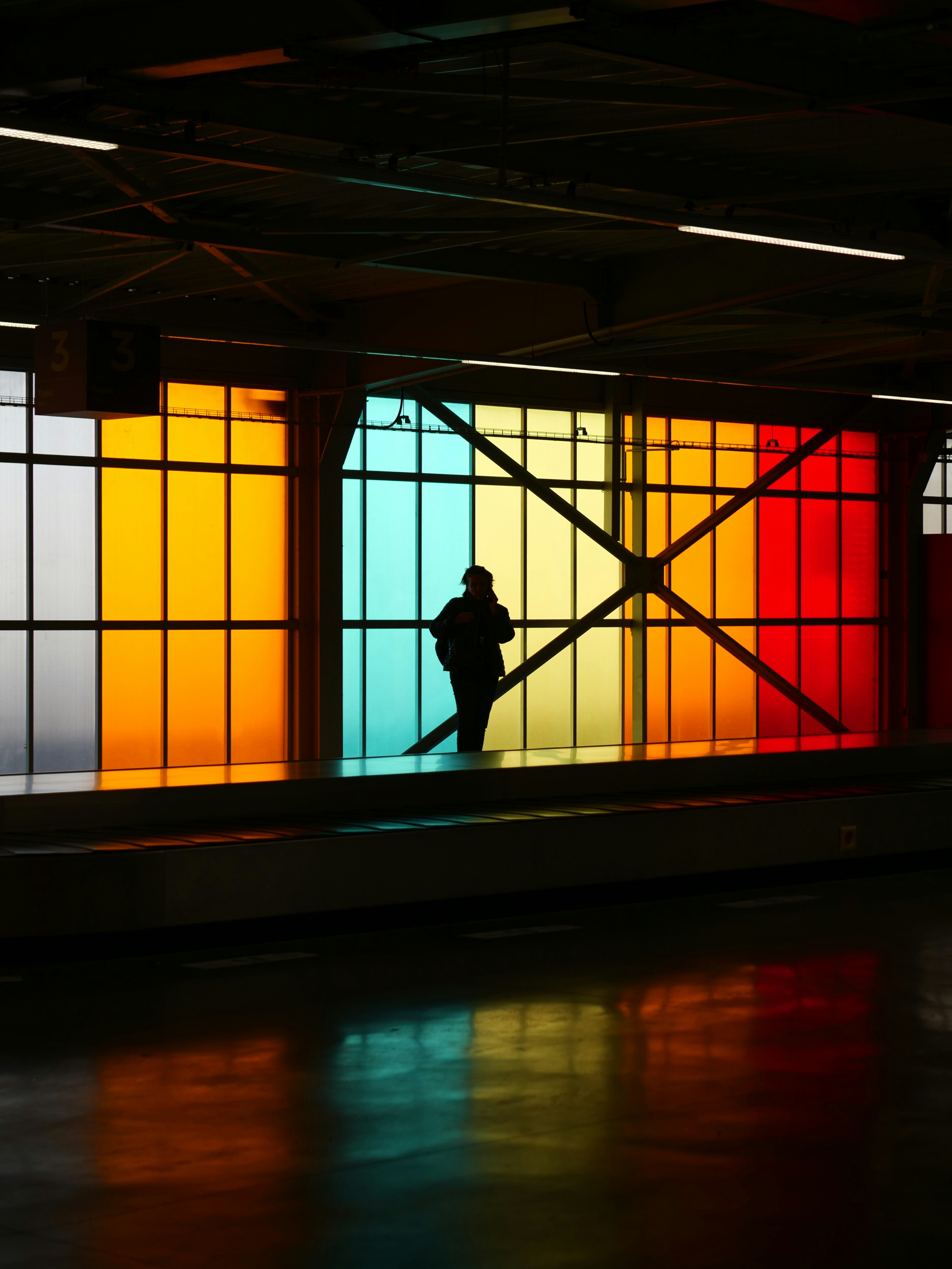 A lone silhouette stands on a quiet platform in front of a backlit, multicolored glass wall. The vibrant panels form a luminous grid that emphasizes the urban transit setting.
