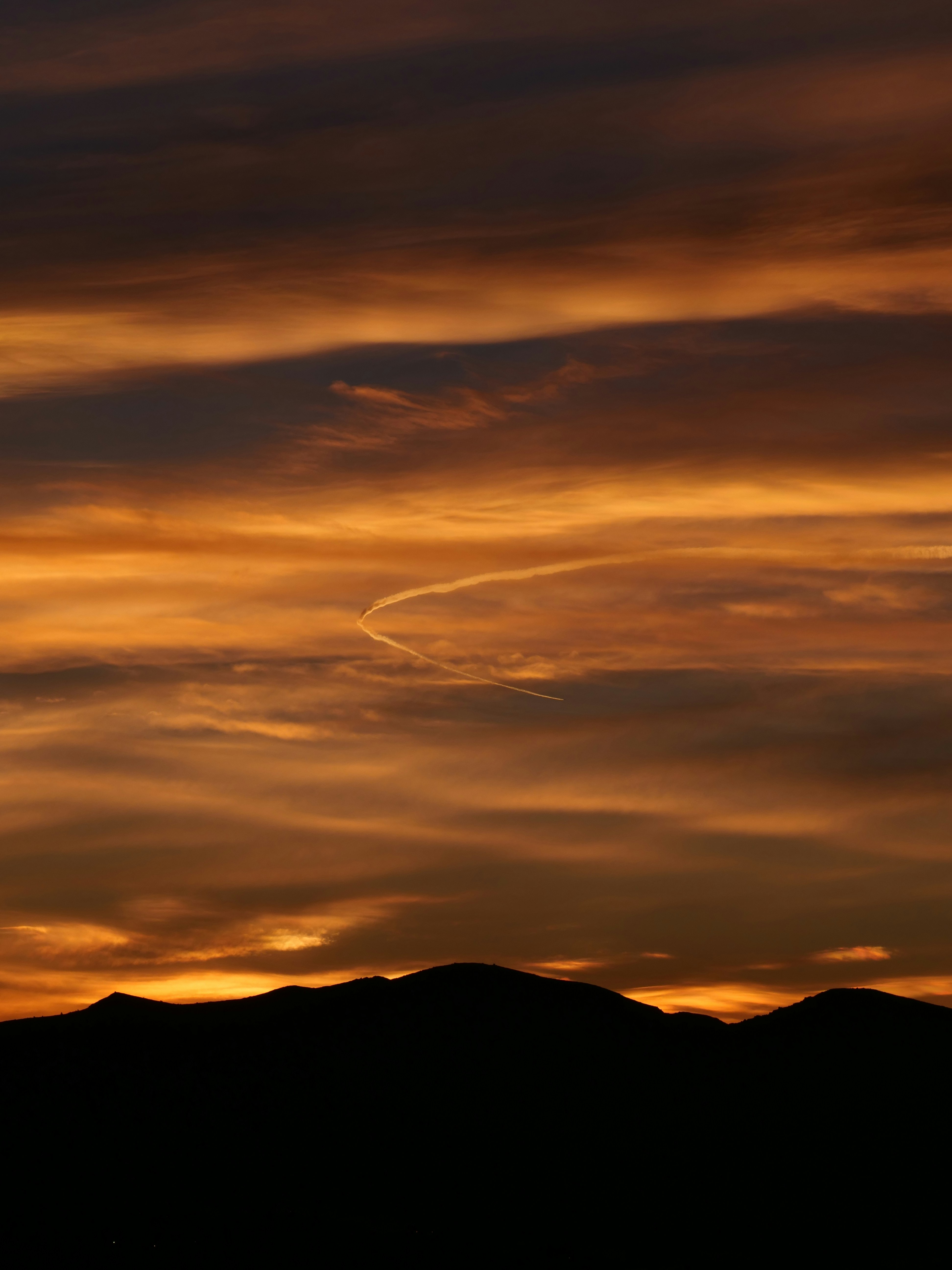 Arc of Ember: A Curved Contrail Over a Silhouetted Ridge at Dusk