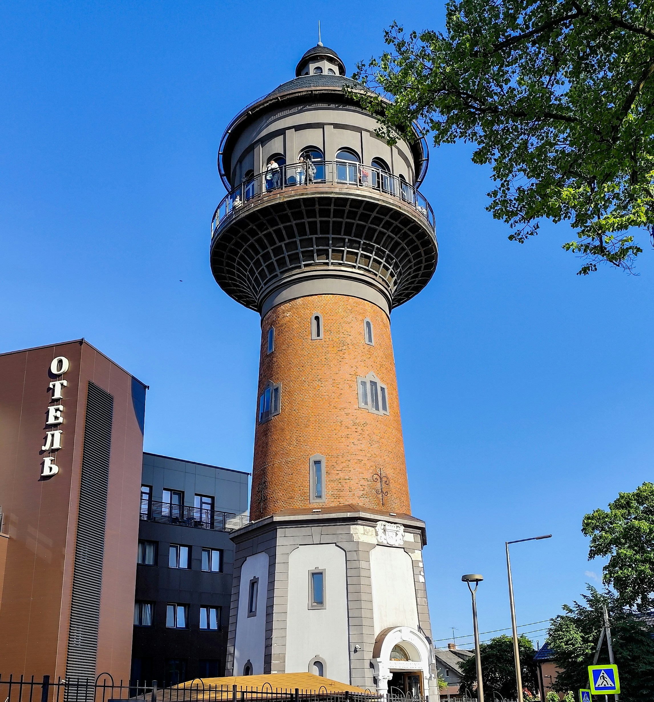 Tall brick observation tower with a circular upper deck rises beside a modern building, set against a clear blue sky and green trees.