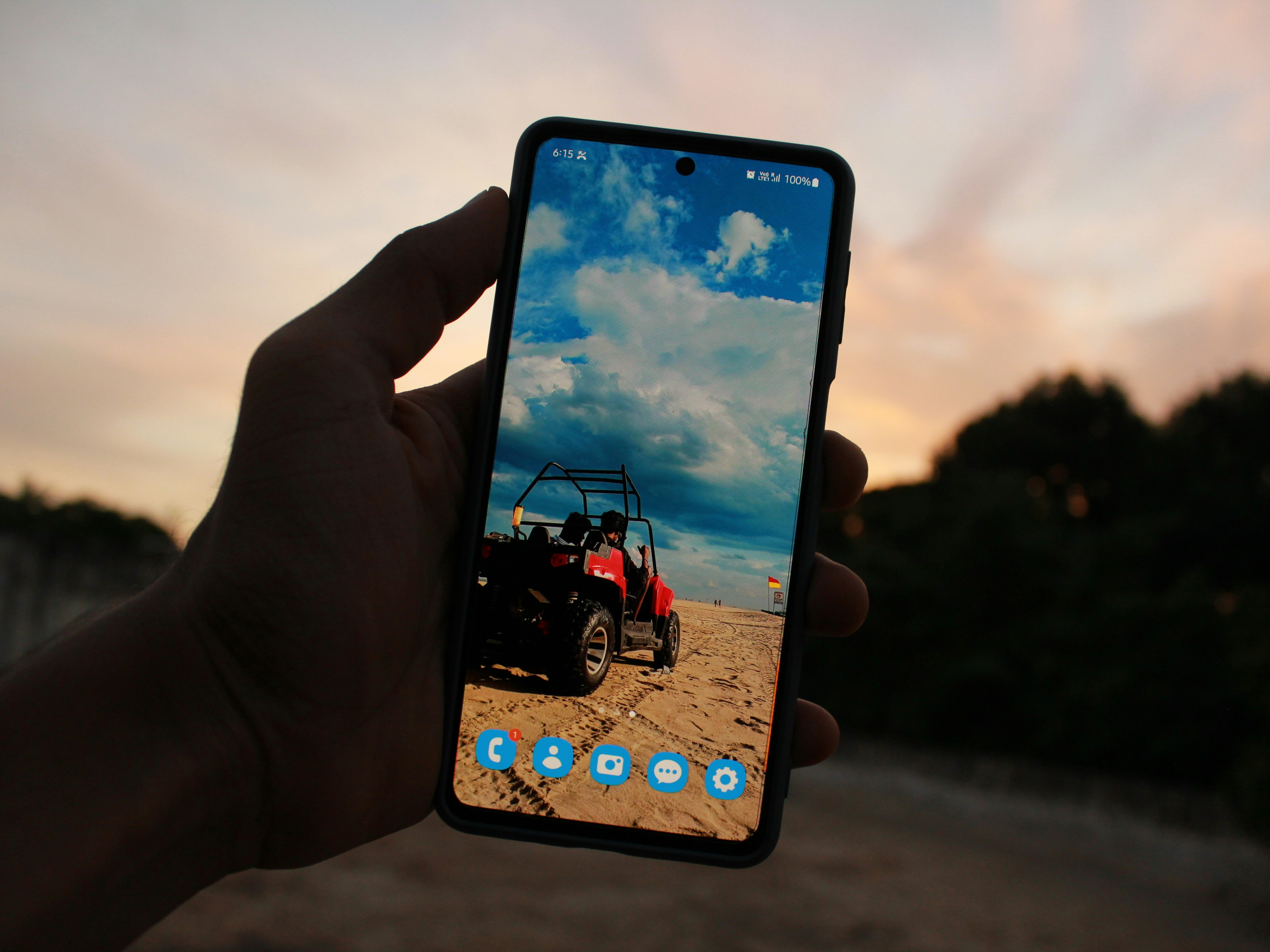 Hand holding a smartphone displaying a vibrant beach scene against a dusky sunset, creating a contrast between digital clarity and natural warmth.