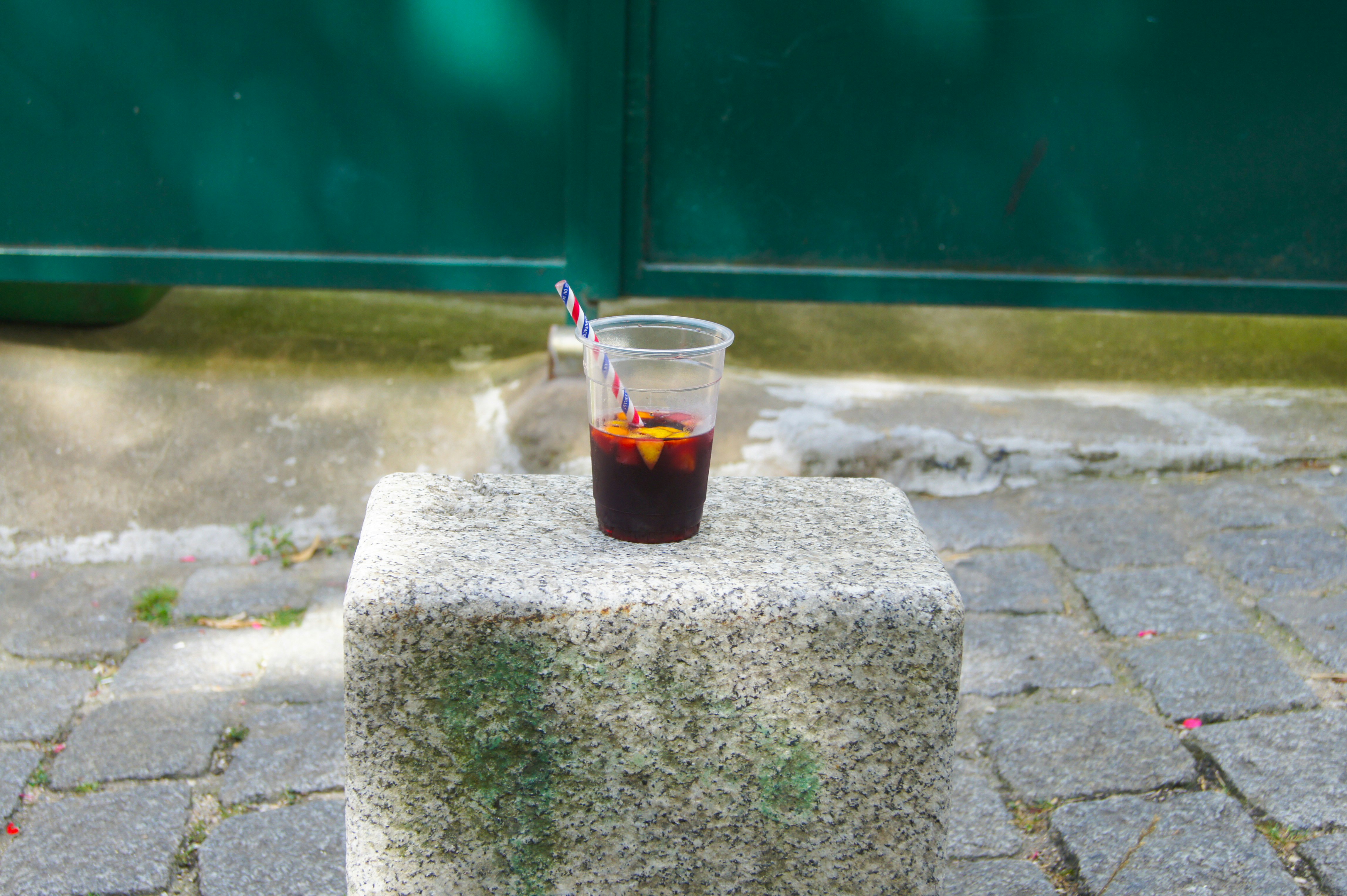 Plastic cup of dark cola with ice and a striped straw sits on a rough granite block in a cobblestone courtyard. A green gate blurs softly in the background.