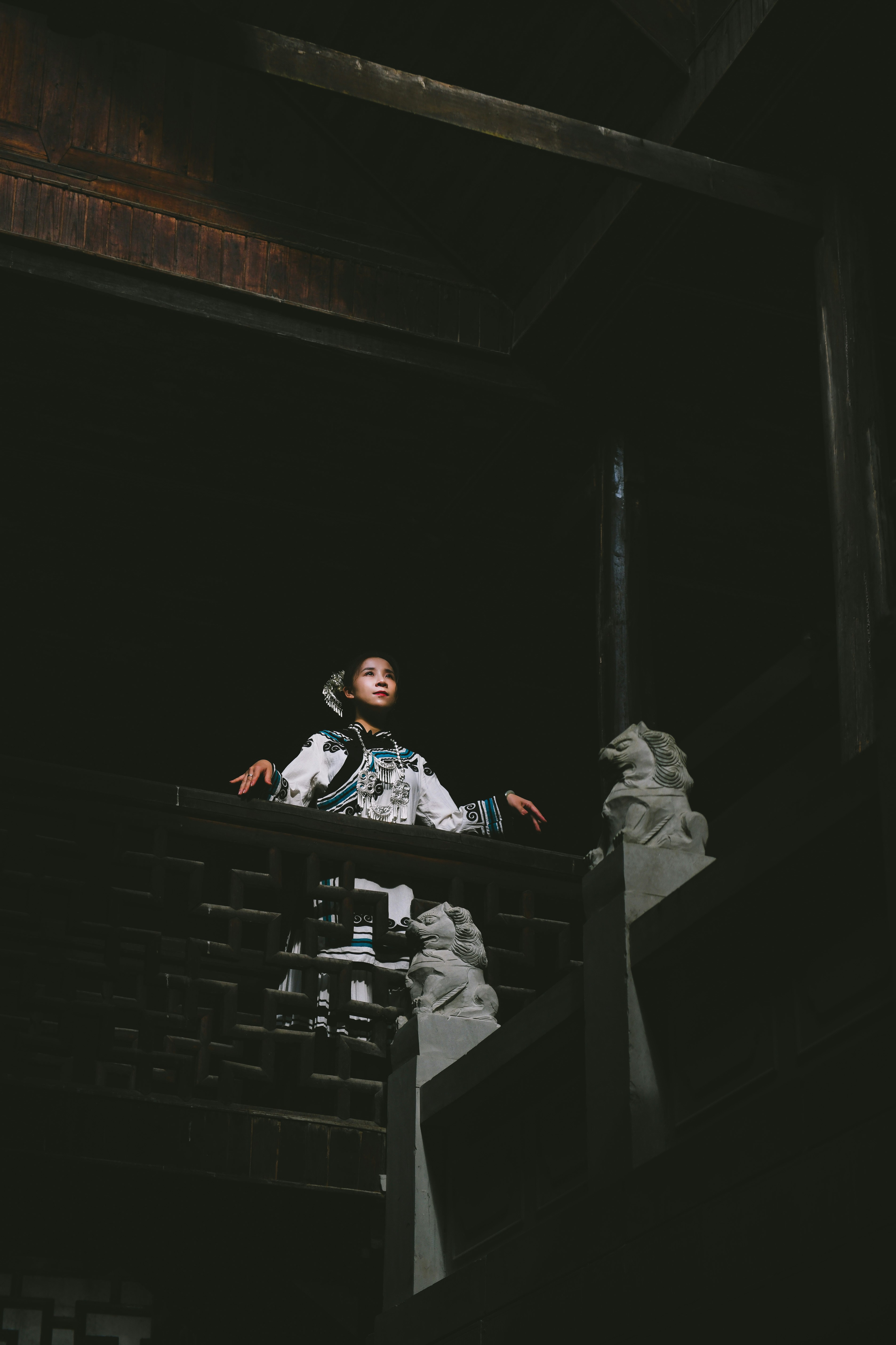 A young woman in traditional attire stands on a balcony, framed by dark wooden architecture and stone lion sculptures. The interplay of light and shadow enhances the scene's mystique.