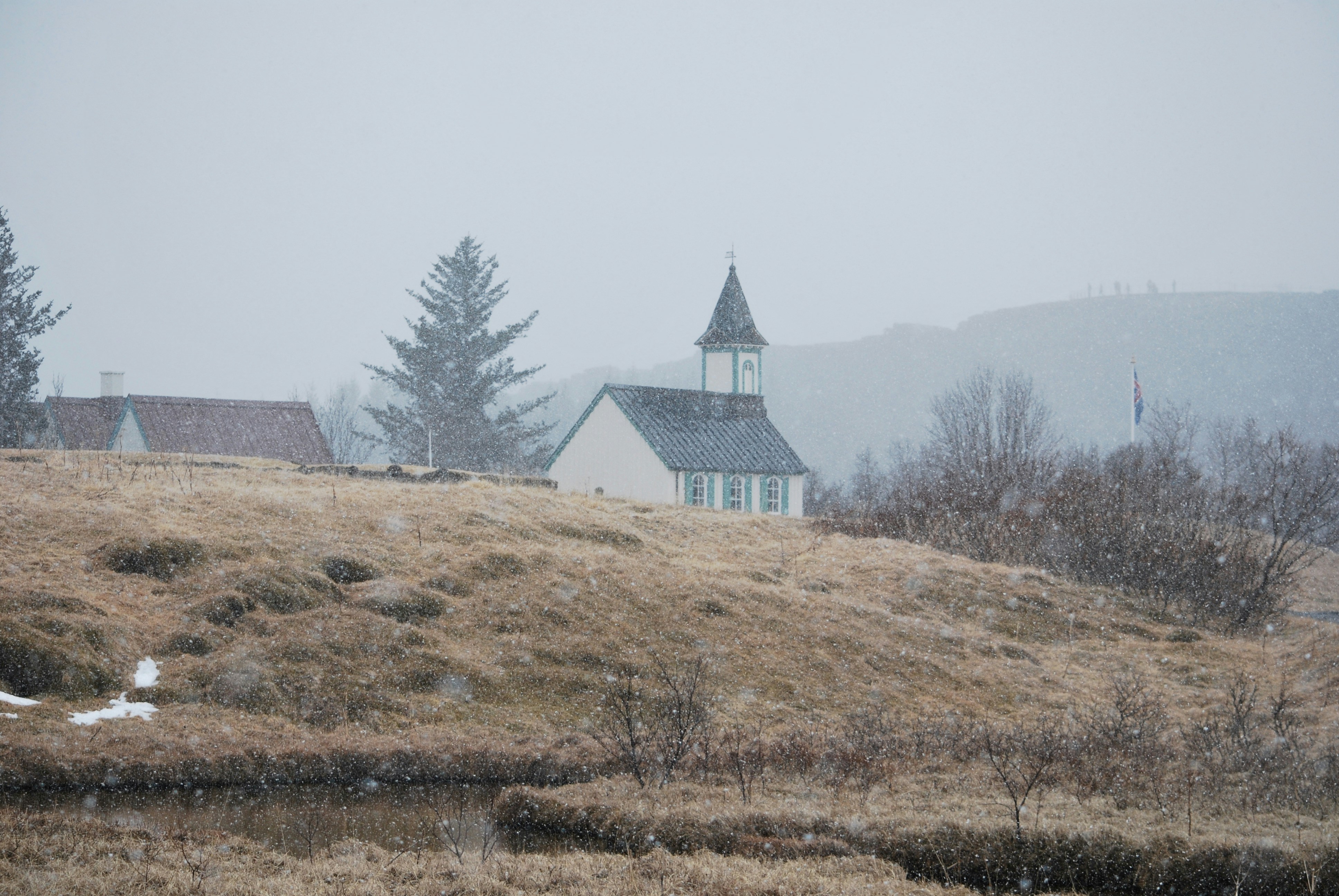 A quaint church stands amidst a snow-covered landscape, surrounded by a muted atmosphere of falling snowflakes and distant hills.