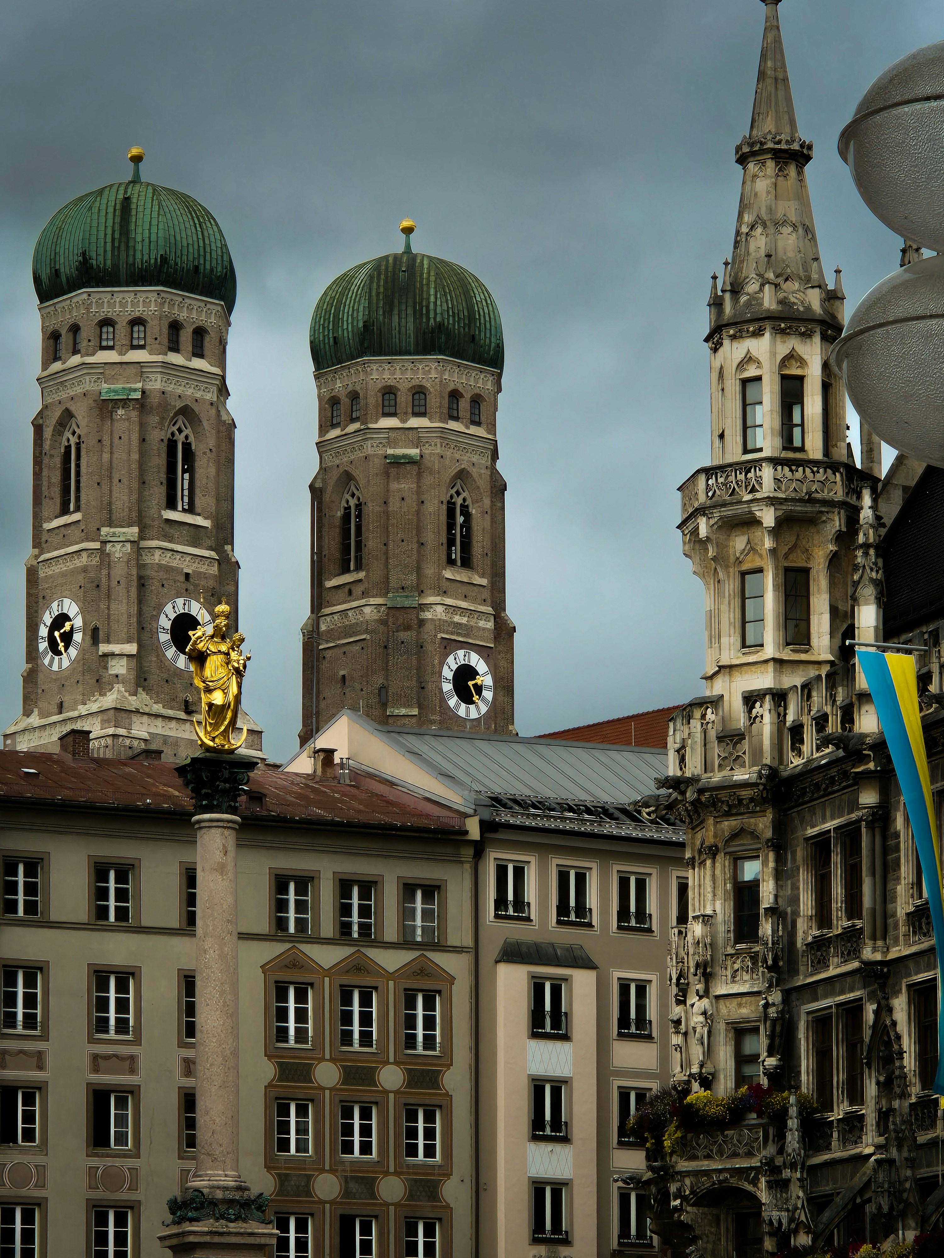 A gilded statue tops a tall column in a historic square, with twin green-domed towers and ornate façades forming the backdrop.