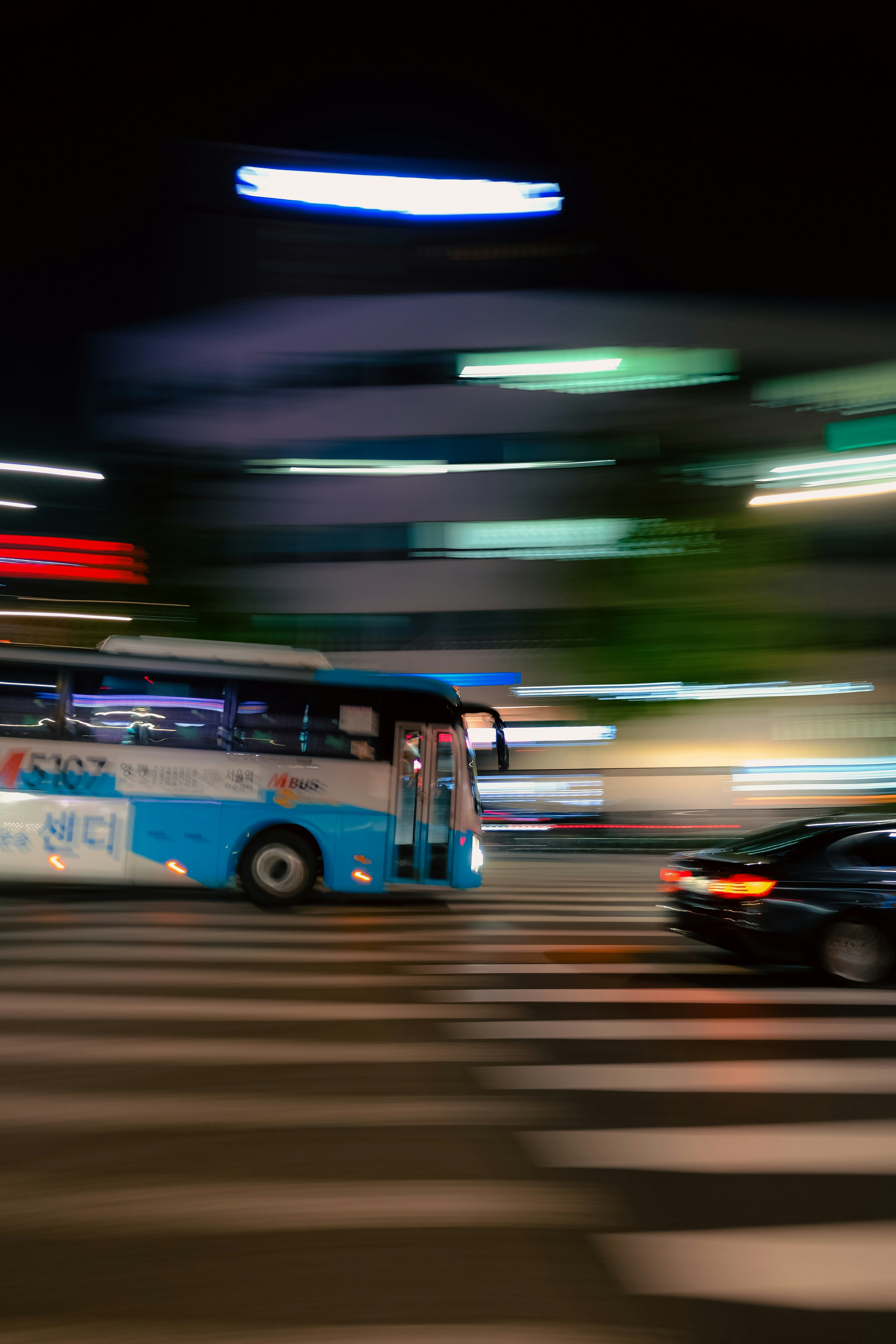A nighttime street scene captured with strong motion blur; a blue city bus moves across a zebra crossing as a black car speeds by, set against streaking neon lights.