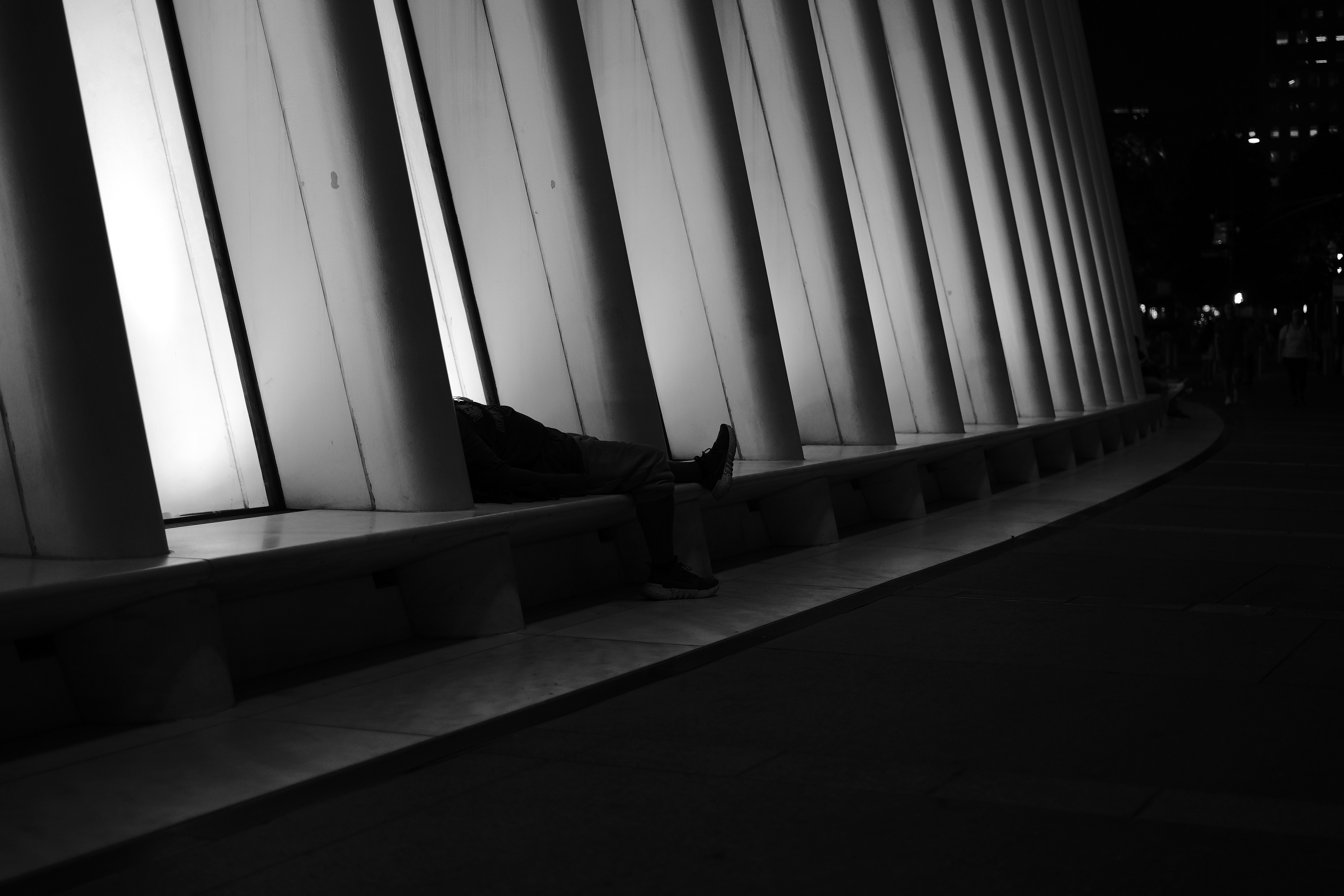 A solitary figure rests against a series of illuminated architectural columns at night, casting intriguing shadows.