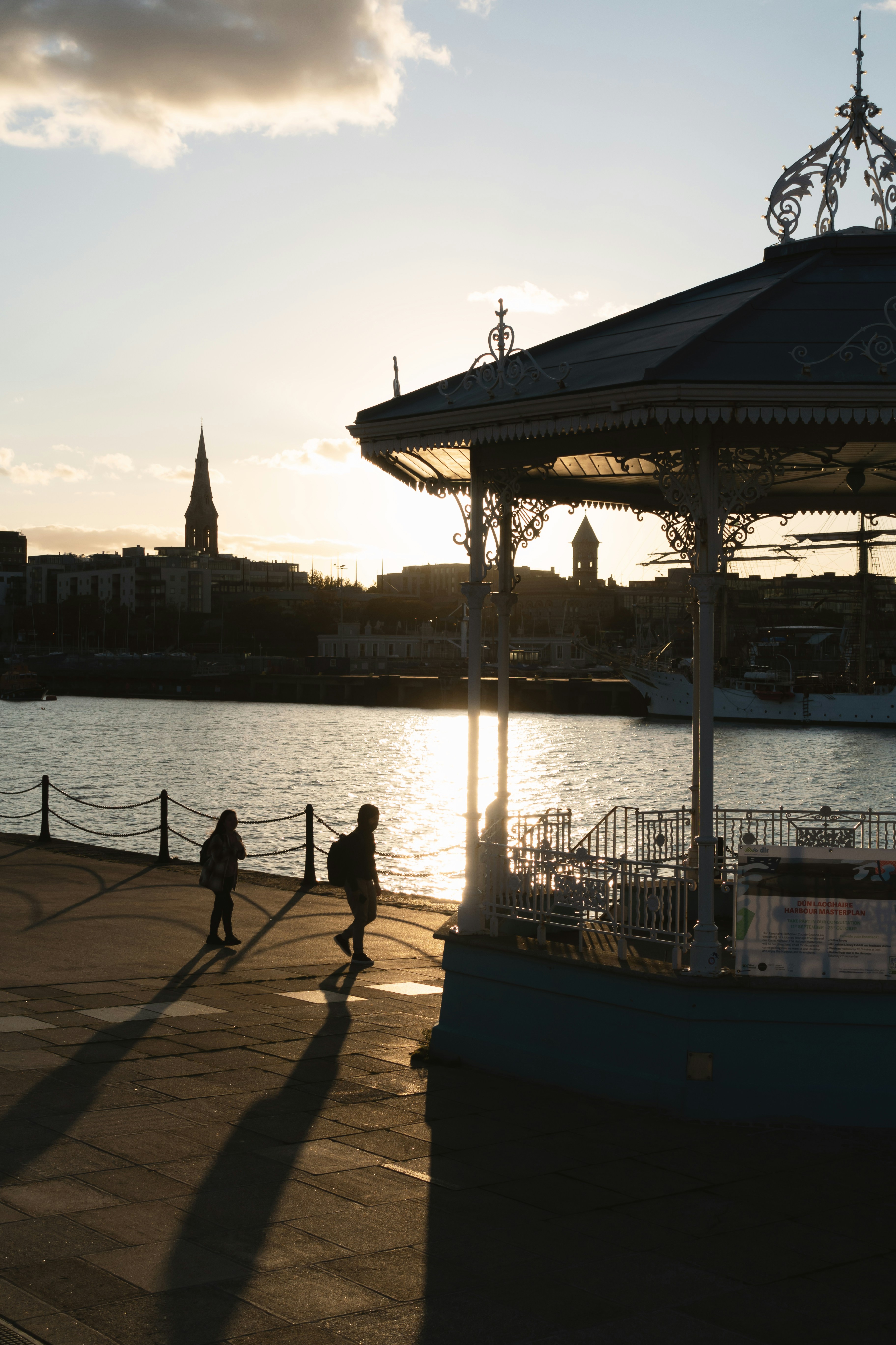 Sunset Promenade: Lacework Gazebo and Shadowed Figures by the Harbor