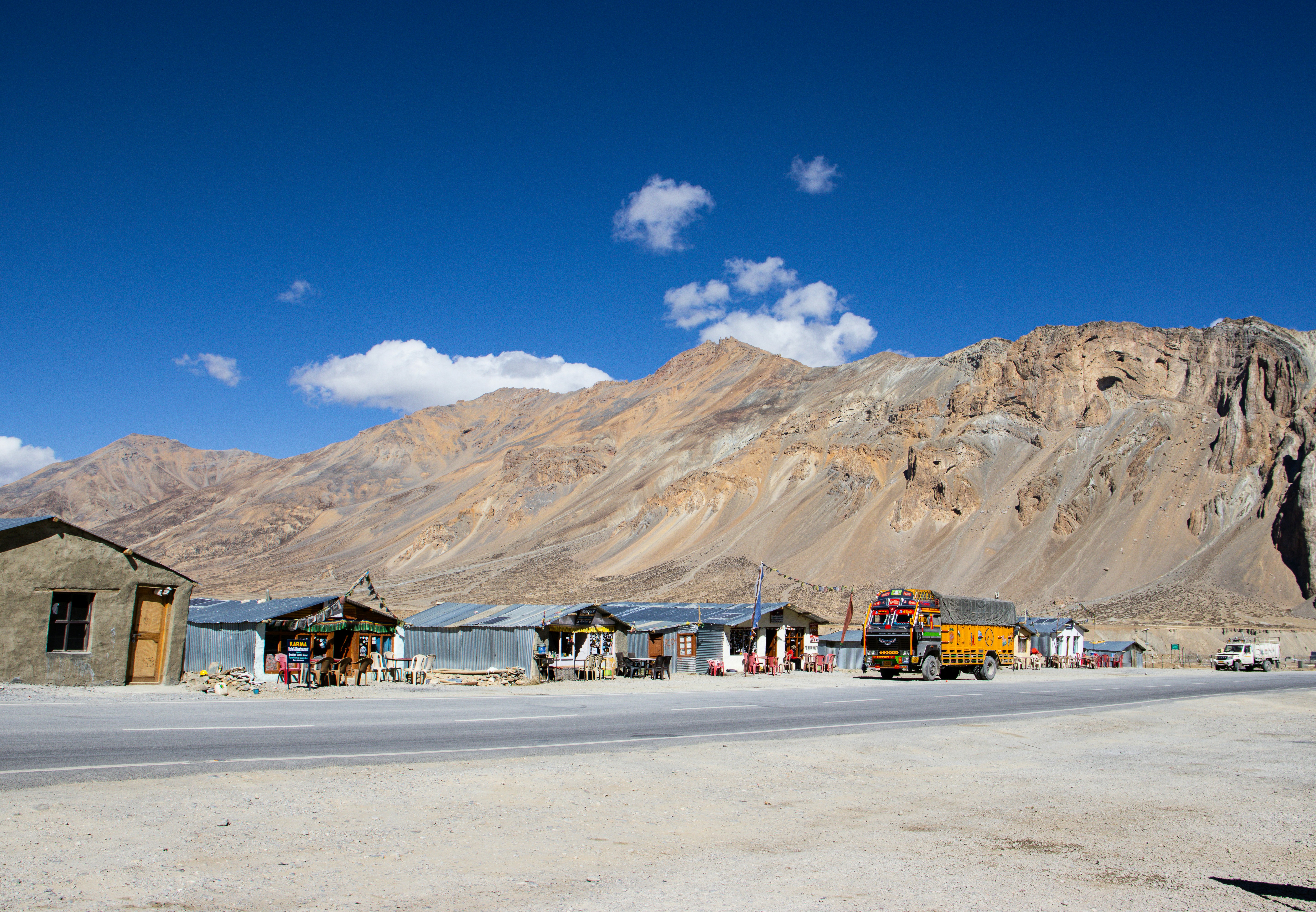 Desert Roadside Horizon: A Bright Truck Among Humble Huts Beneath Monumental Peaks
