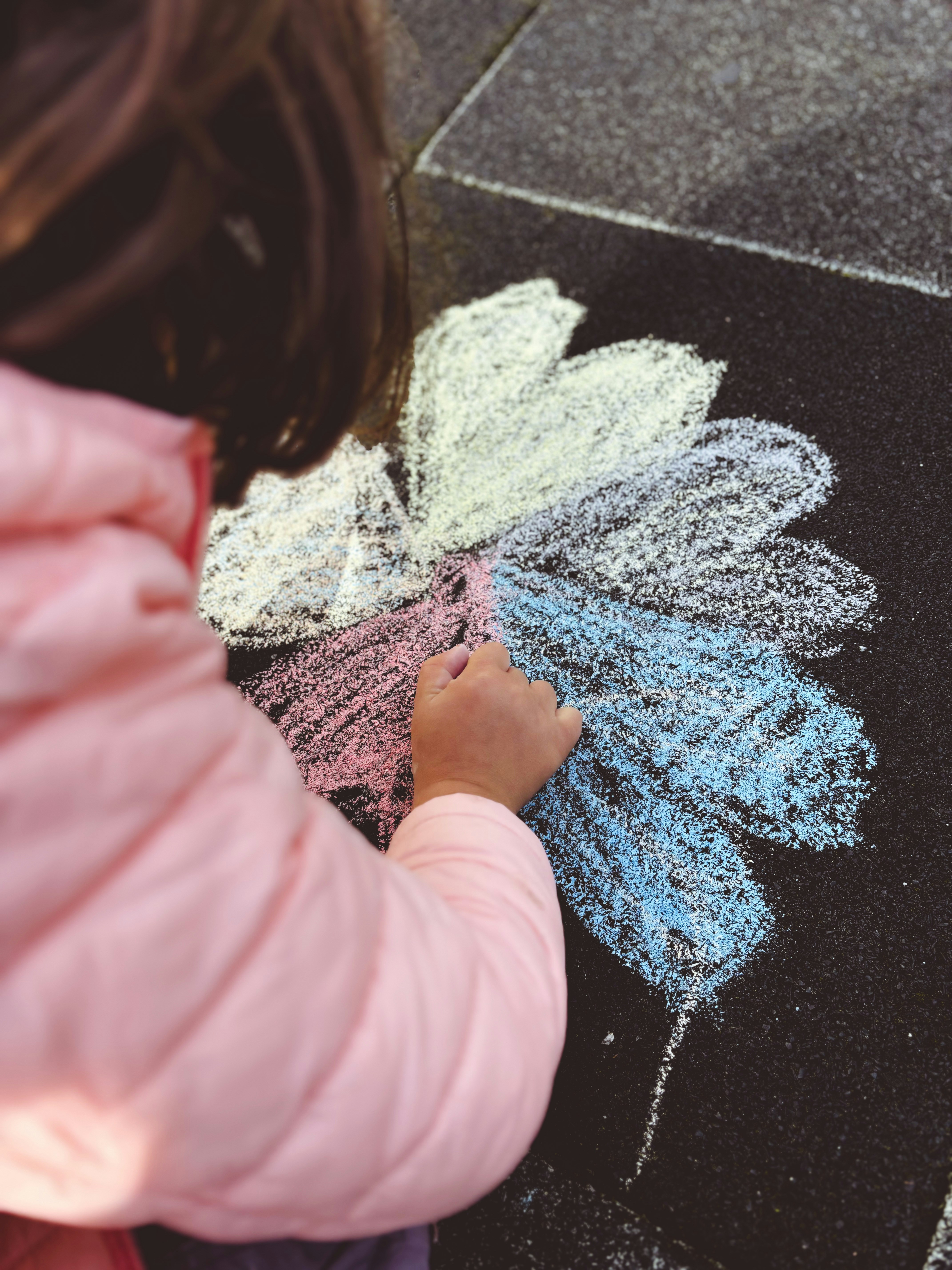 A child in a pink jacket draws a large pastel-colored flower with sidewalk chalk on dark asphalt. The photo highlights the hand at the bloom's center and the chalk dust texture on the pavement.