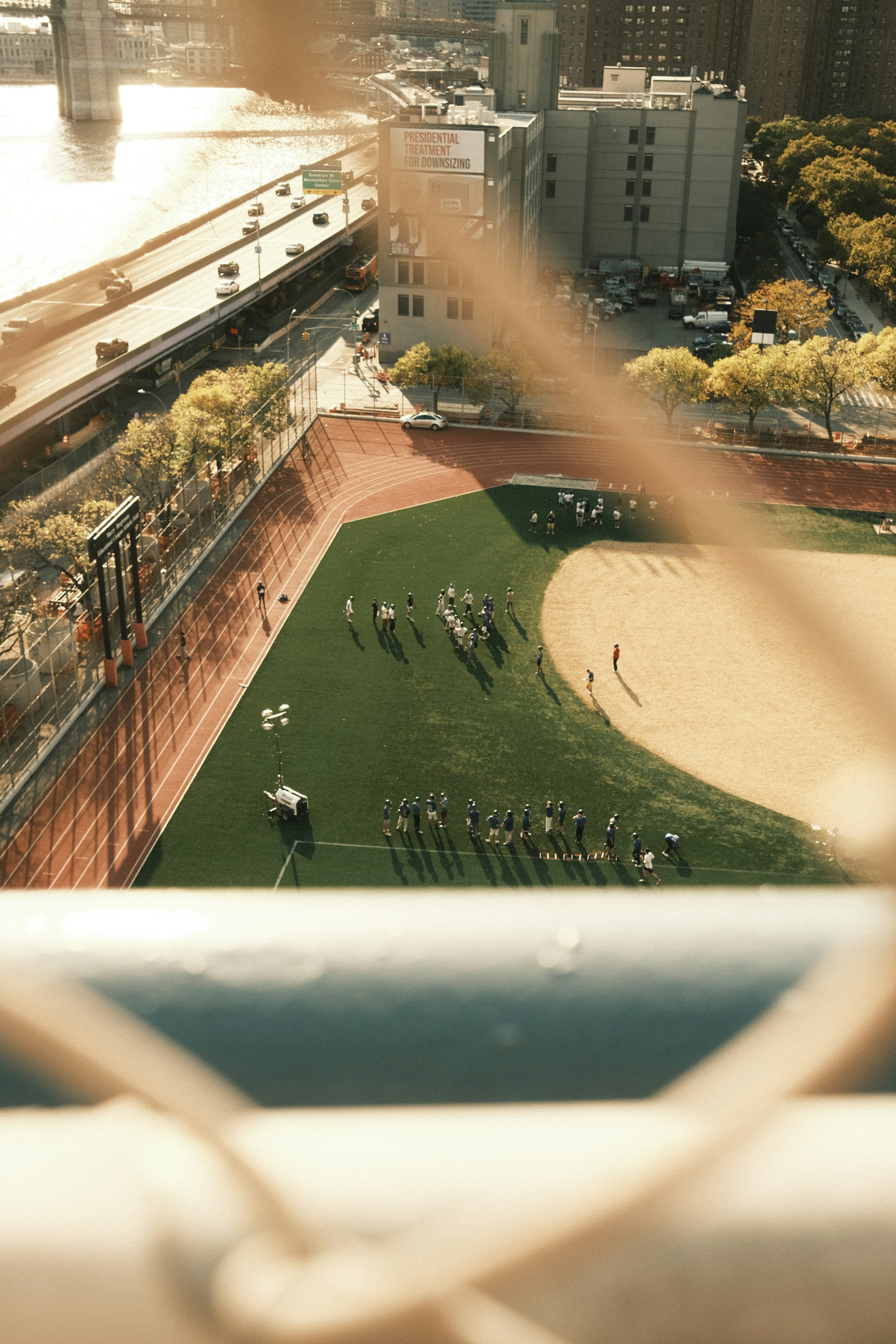 Aerial view of a sports practice on a green field, surrounded by urban architecture and autumn foliage. The scene captures players engaged in activity under the warm glow of evening light.