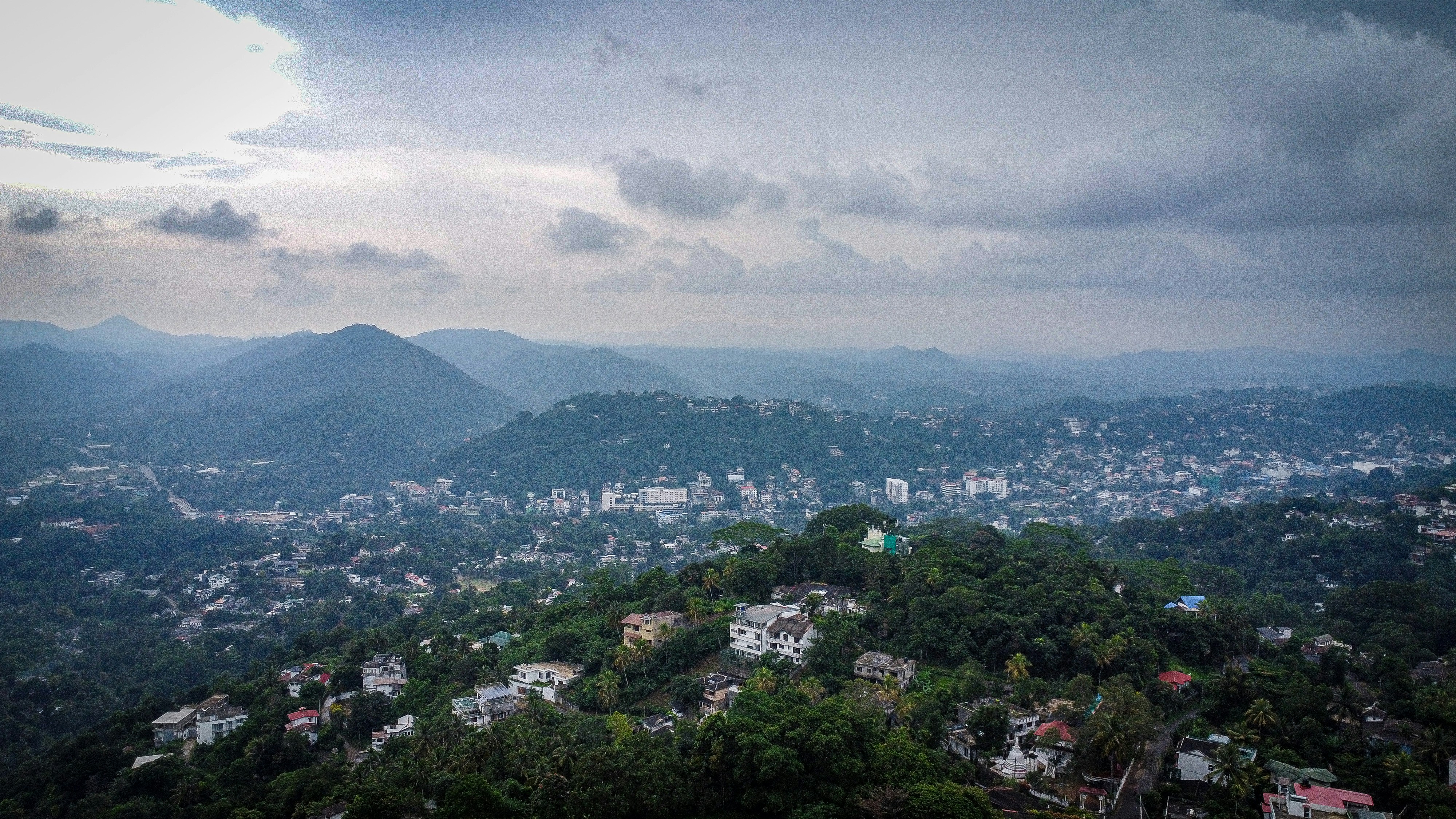Aerial view of a lush hillside community surrounded by distant mountains under a cloudy sky. The scene captures the blend of nature and urban life.