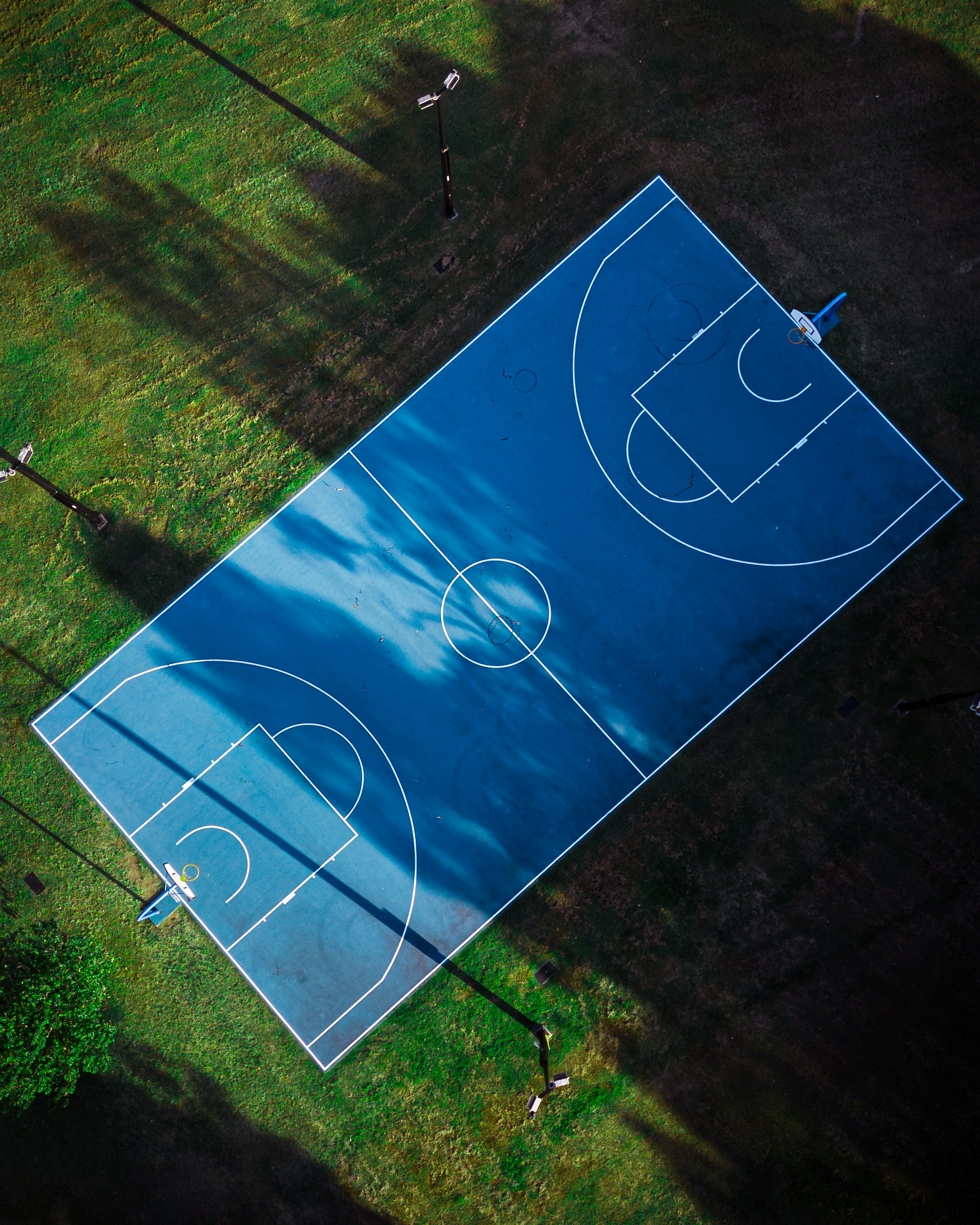 Drone photograph of a blue outdoor basketball court with white lines on green grass, shadows and trees around.