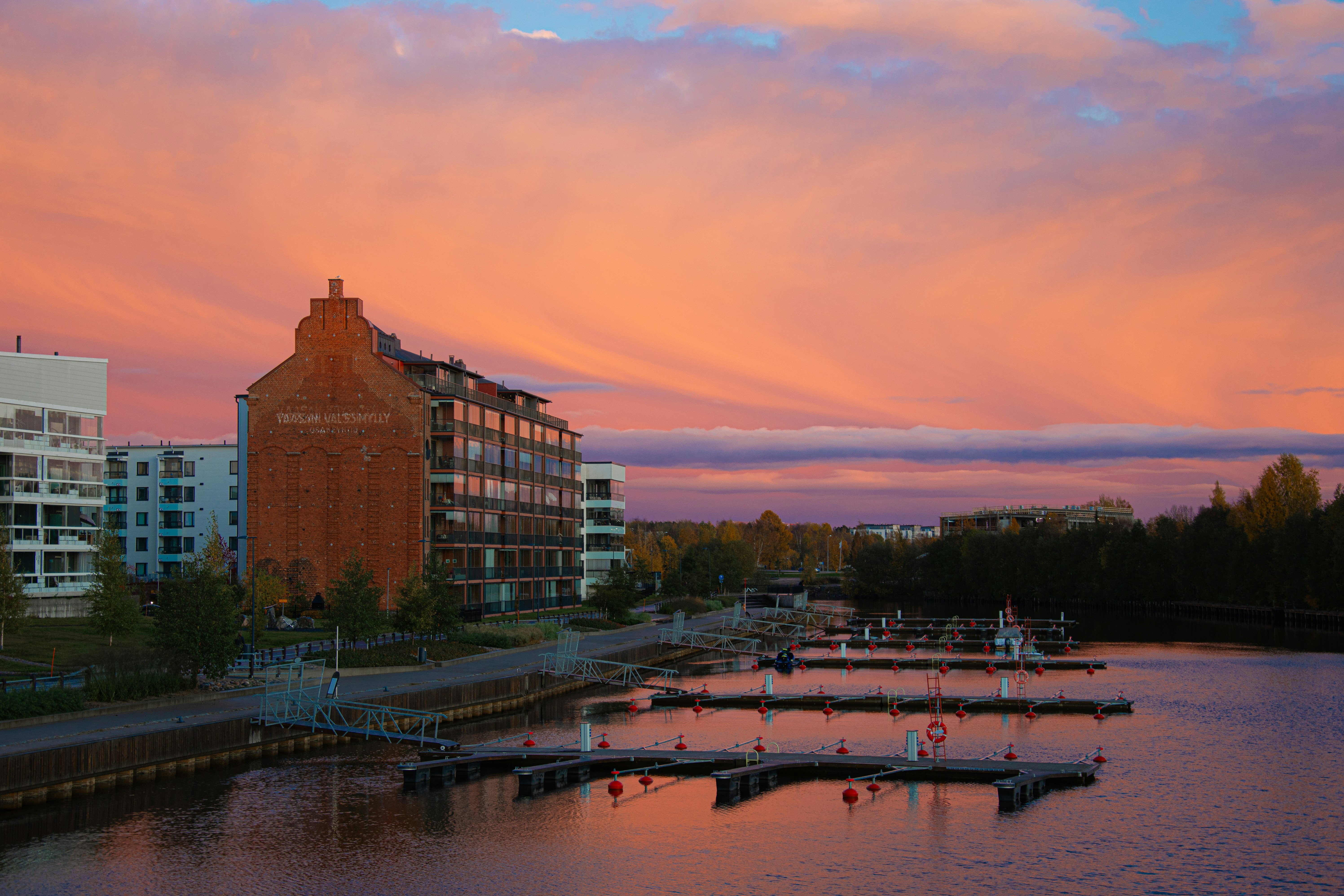 Vibrant sunset sky over riverside buildings and empty docks, reflecting warm hues on calm water.