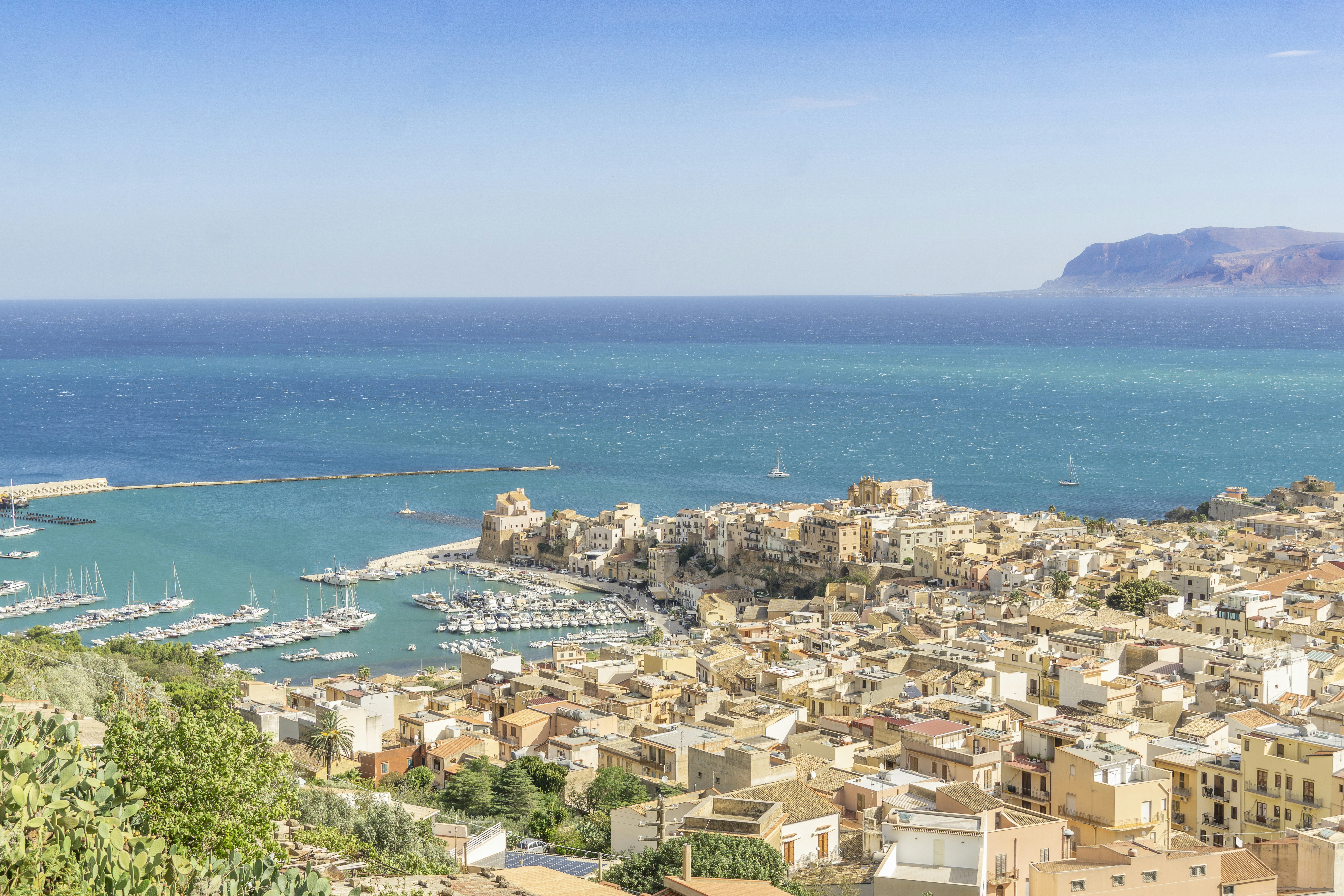 Scenic view of a Mediterranean town nestled by a turquoise sea with a bustling marina and distant mountains on the horizon.