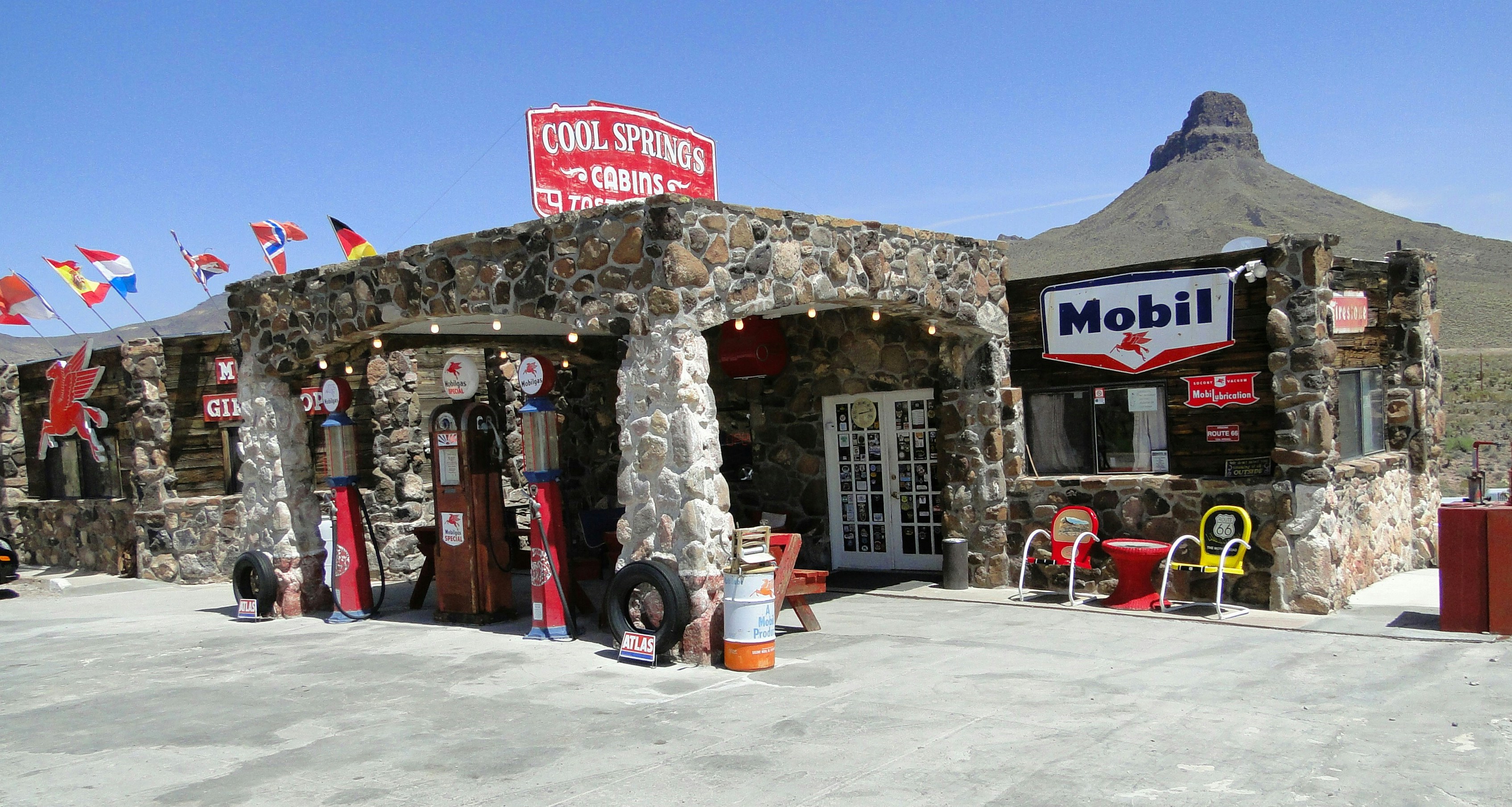 Stone-walled desert service stop with banners, vintage chairs, and a Mobil sign. A distant cone-shaped hill rises against a clear blue sky.