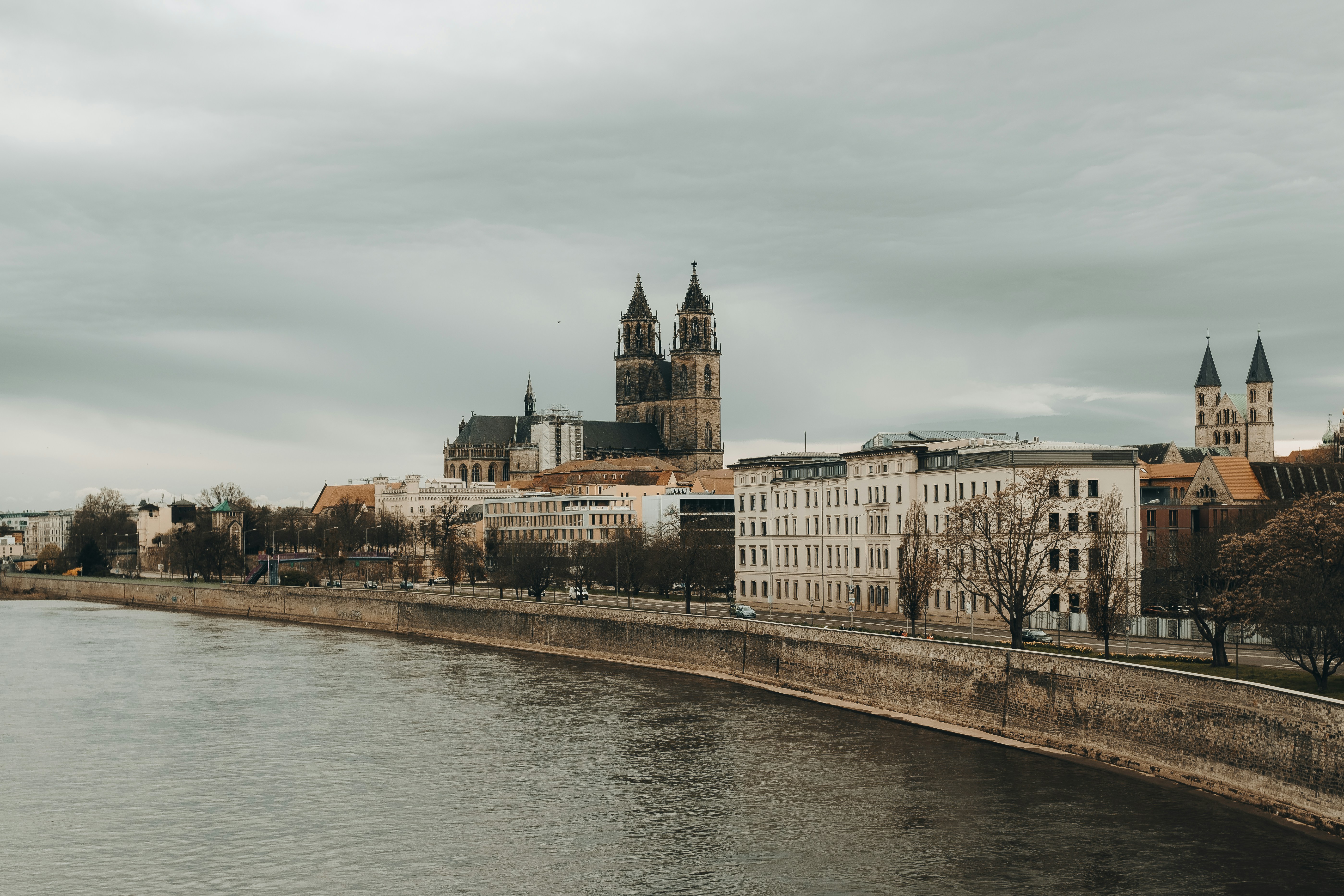 Historic cathedral and waterfront buildings along a riverbank under cloudy skies.