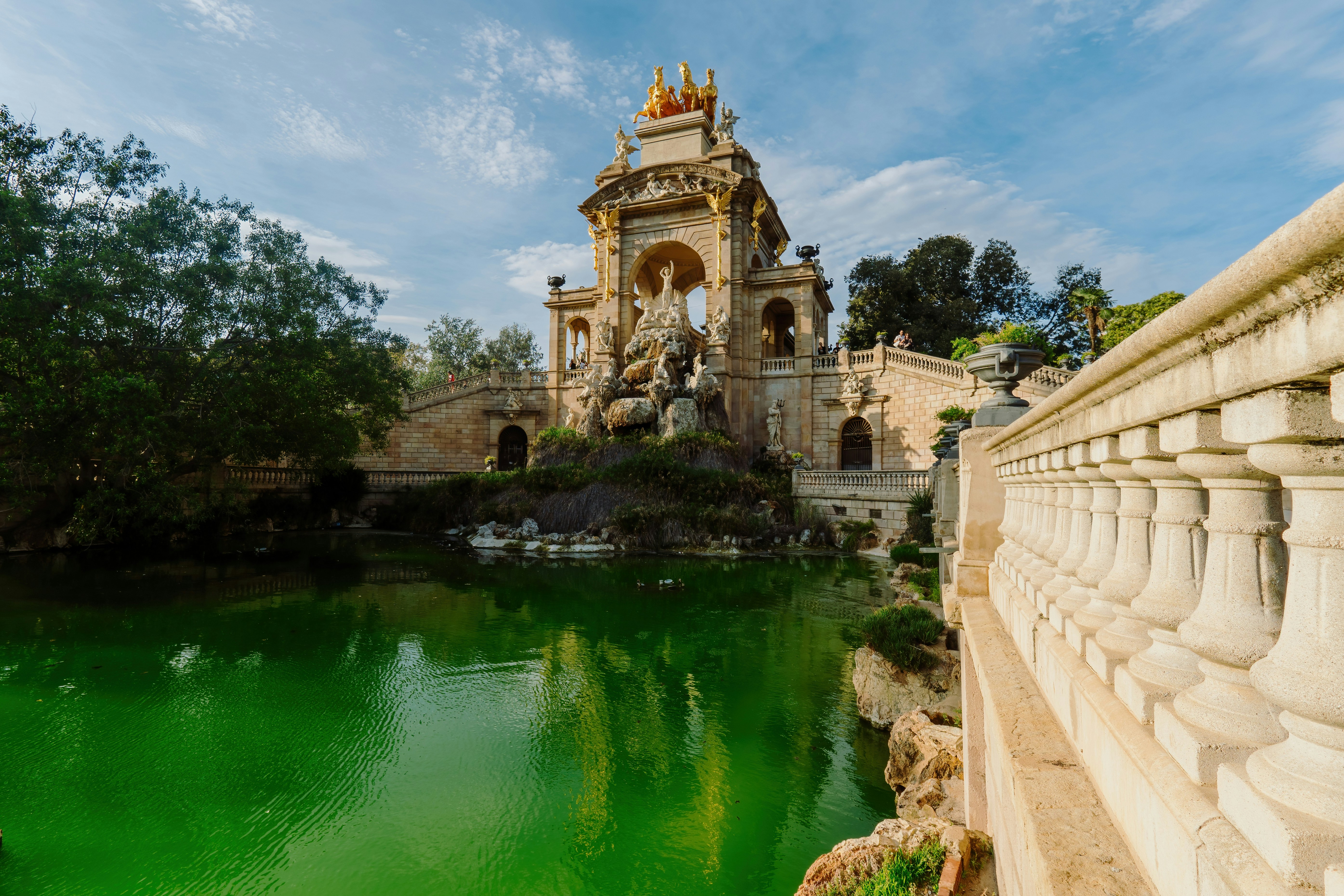 Historic fountain structure with intricate sculptures beside a vibrant green pond under a blue sky.