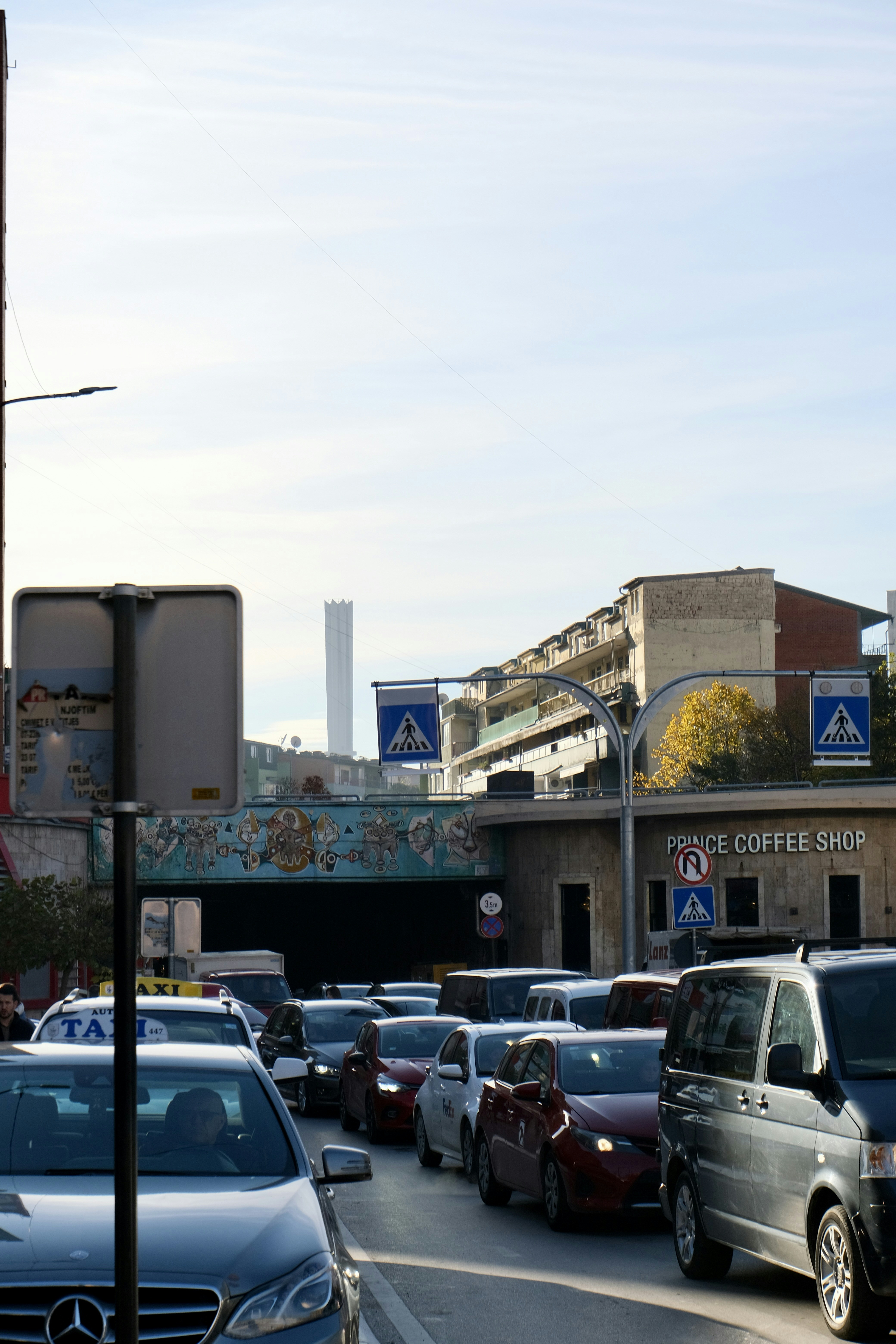Traffic jam under a mural-adorned bridge near a coffee shop on a sunny day.