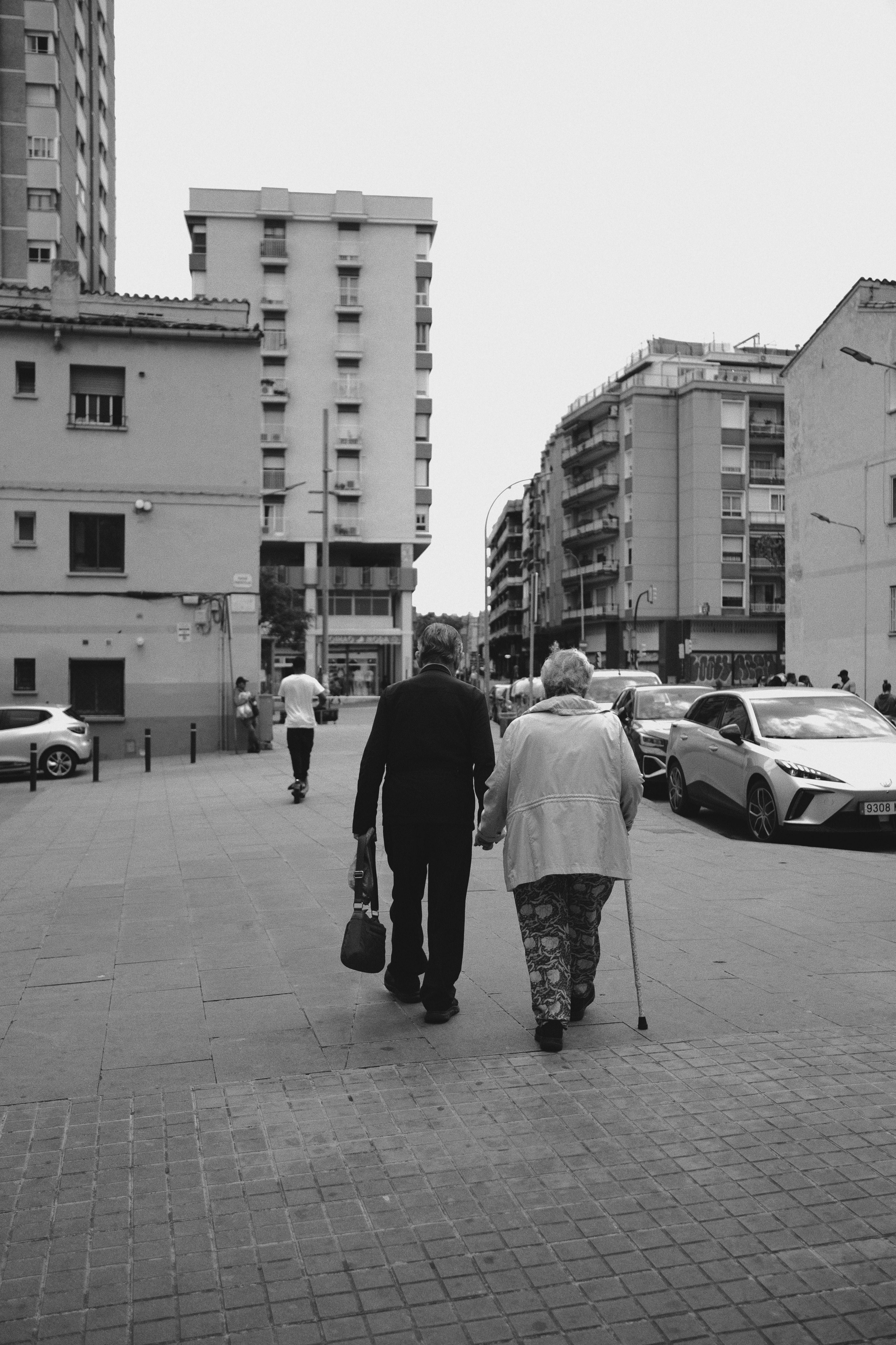 Two elderly individuals walking hand-in-hand down a city street, surrounded by modern architecture and urban life.