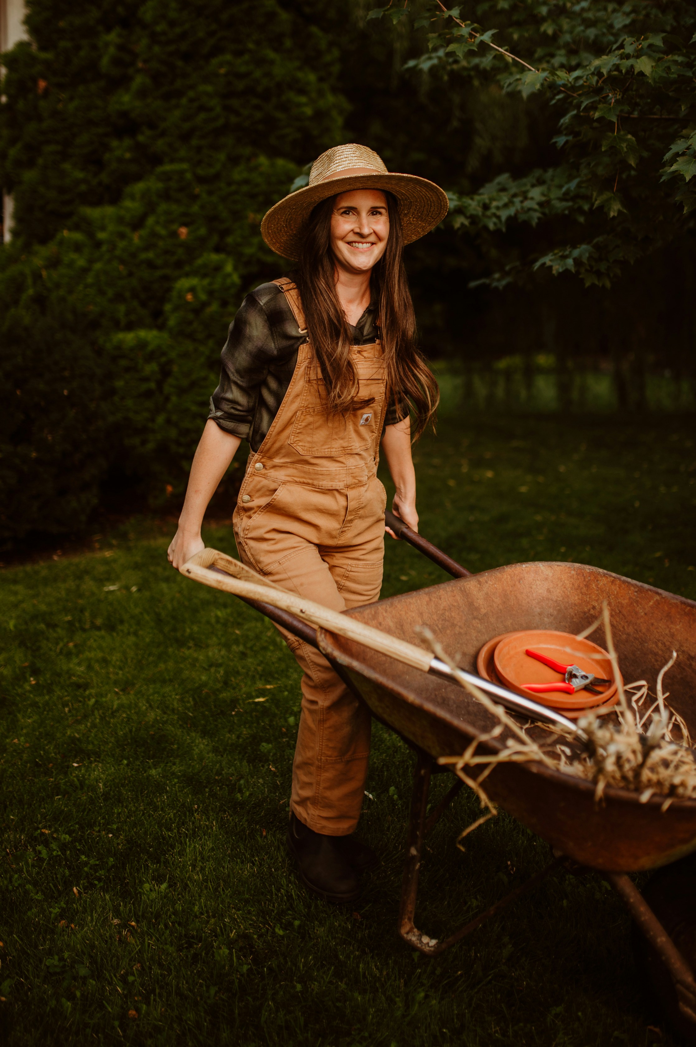 Woman gardening in brown overalls pushing an old metal wheelbarrow accross her lush lawn - yard cleanup, yard work
