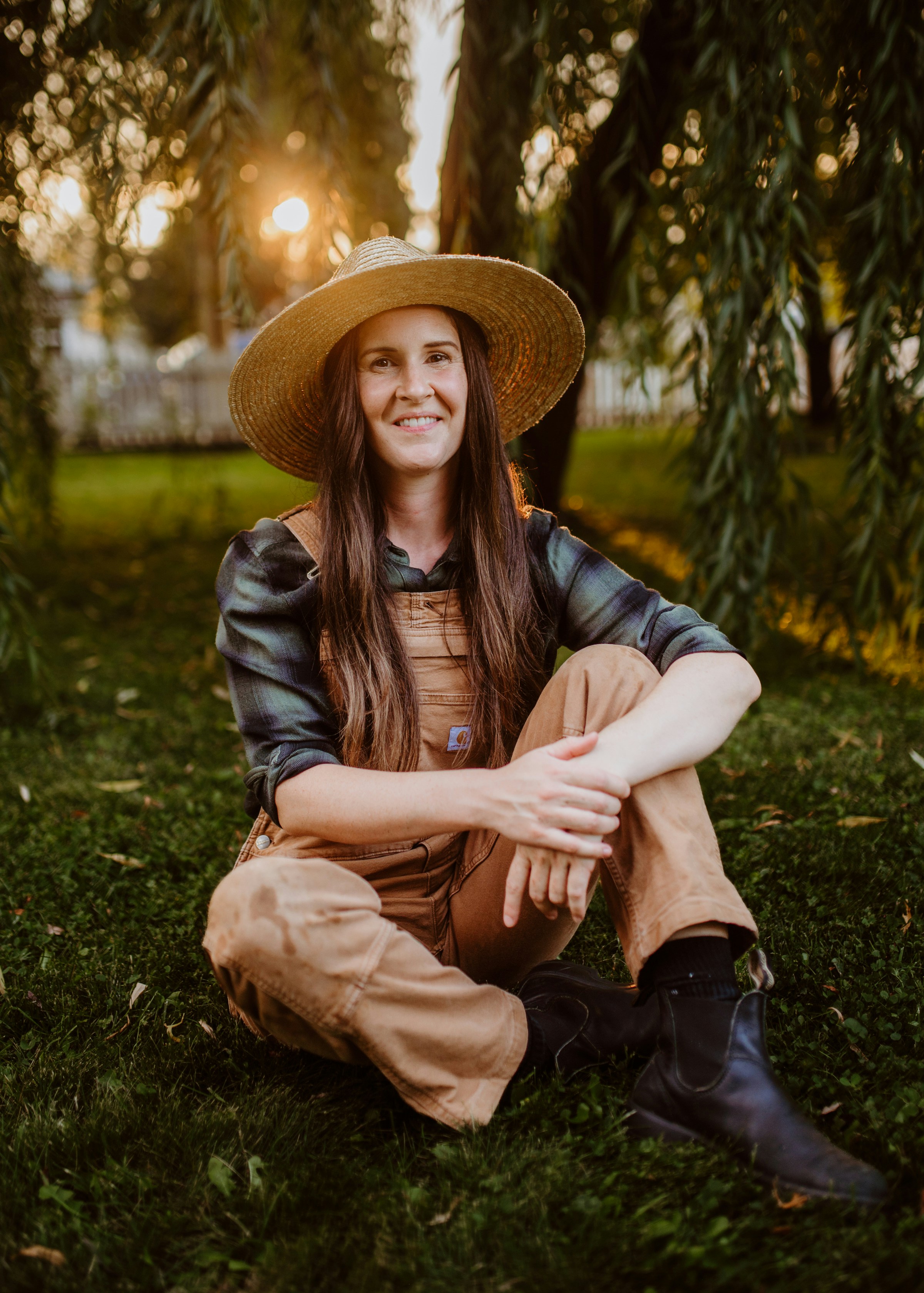 Happy landscaper sitting on grass smiling wearing brown overalls, plaid shirt, and straw hat, by willow tree on lush green lawn | A woman sitting on the ground wearing a hat