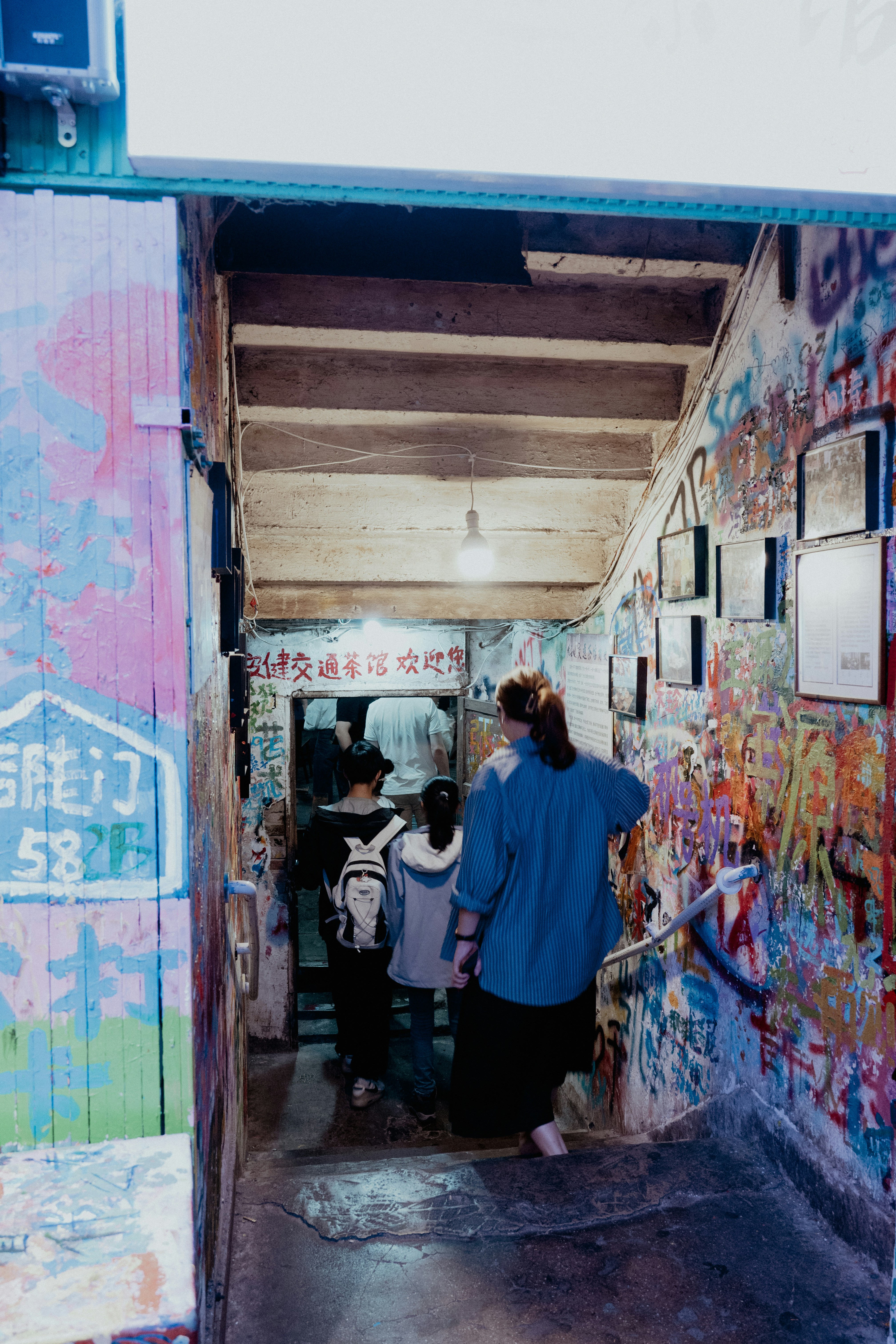 Vibrantly painted walls line a staircase leading into a bustling underground space, with figures ascending and descending. The glow of a hanging light adds warmth to the scene.