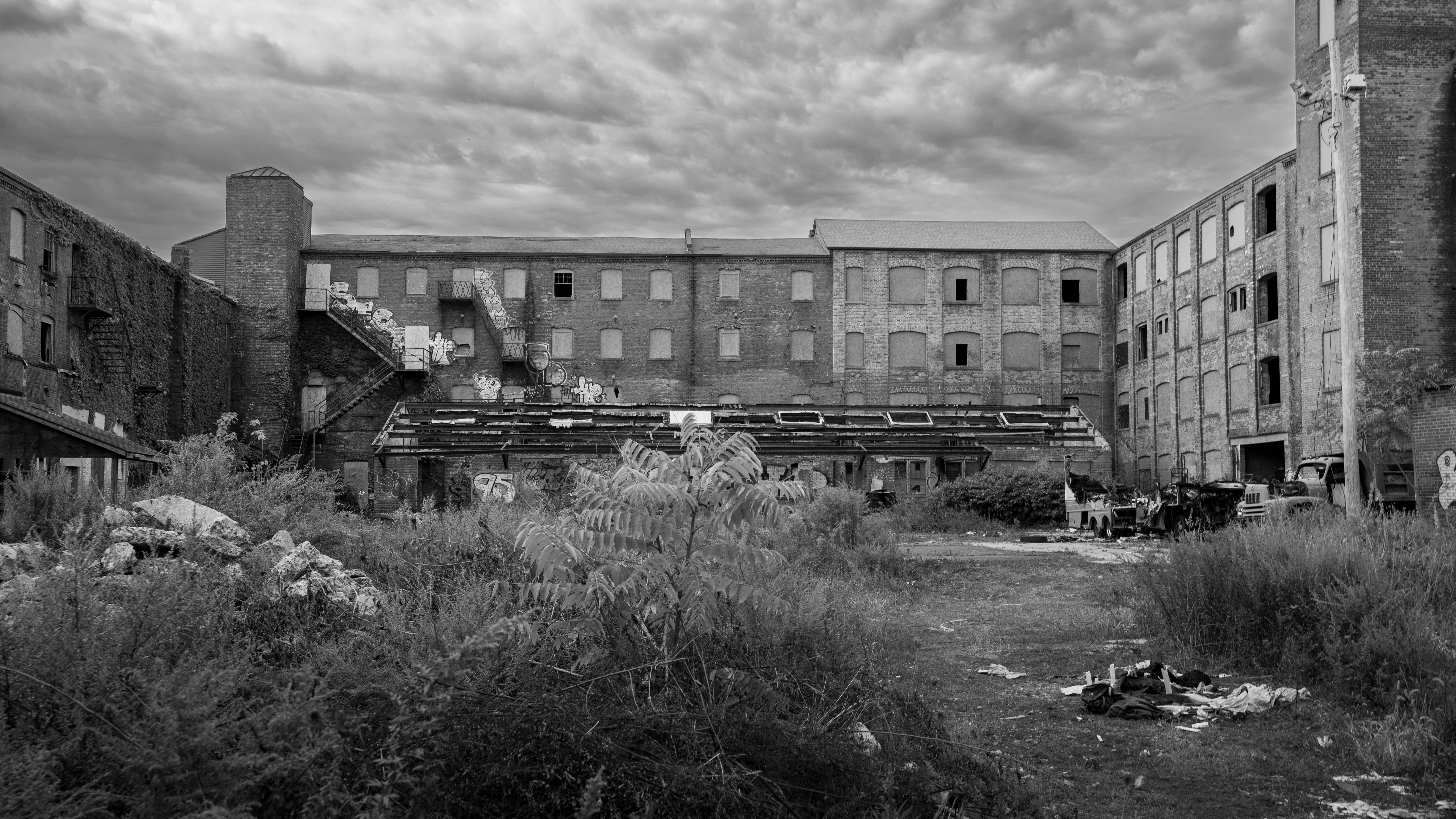 Abandoned brick factory with overgrown vegetation and cloudy sky.