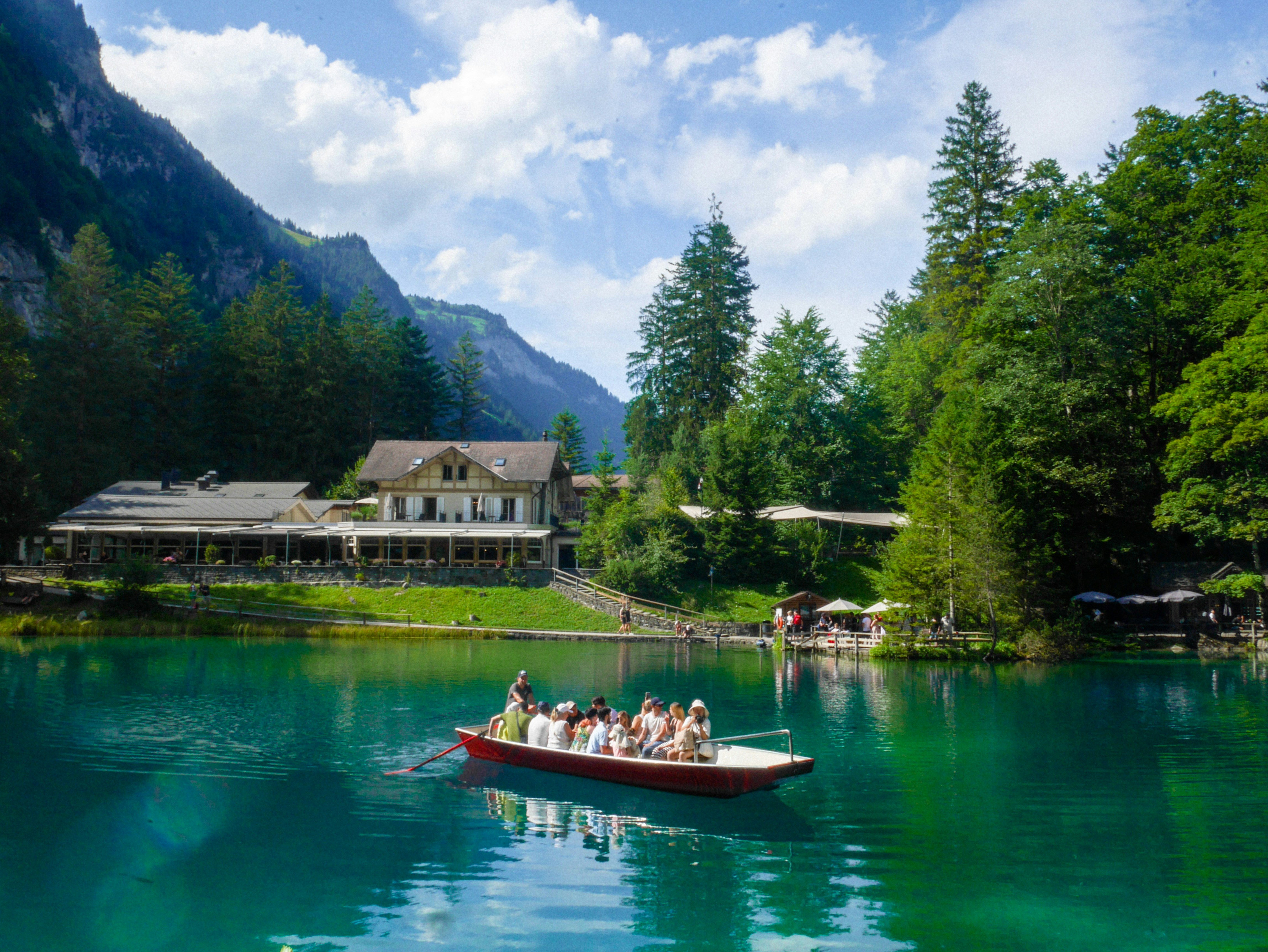 Tour boat glides across a turquoise alpine lake, with a lakeside lodge and forested peaks in the background.
