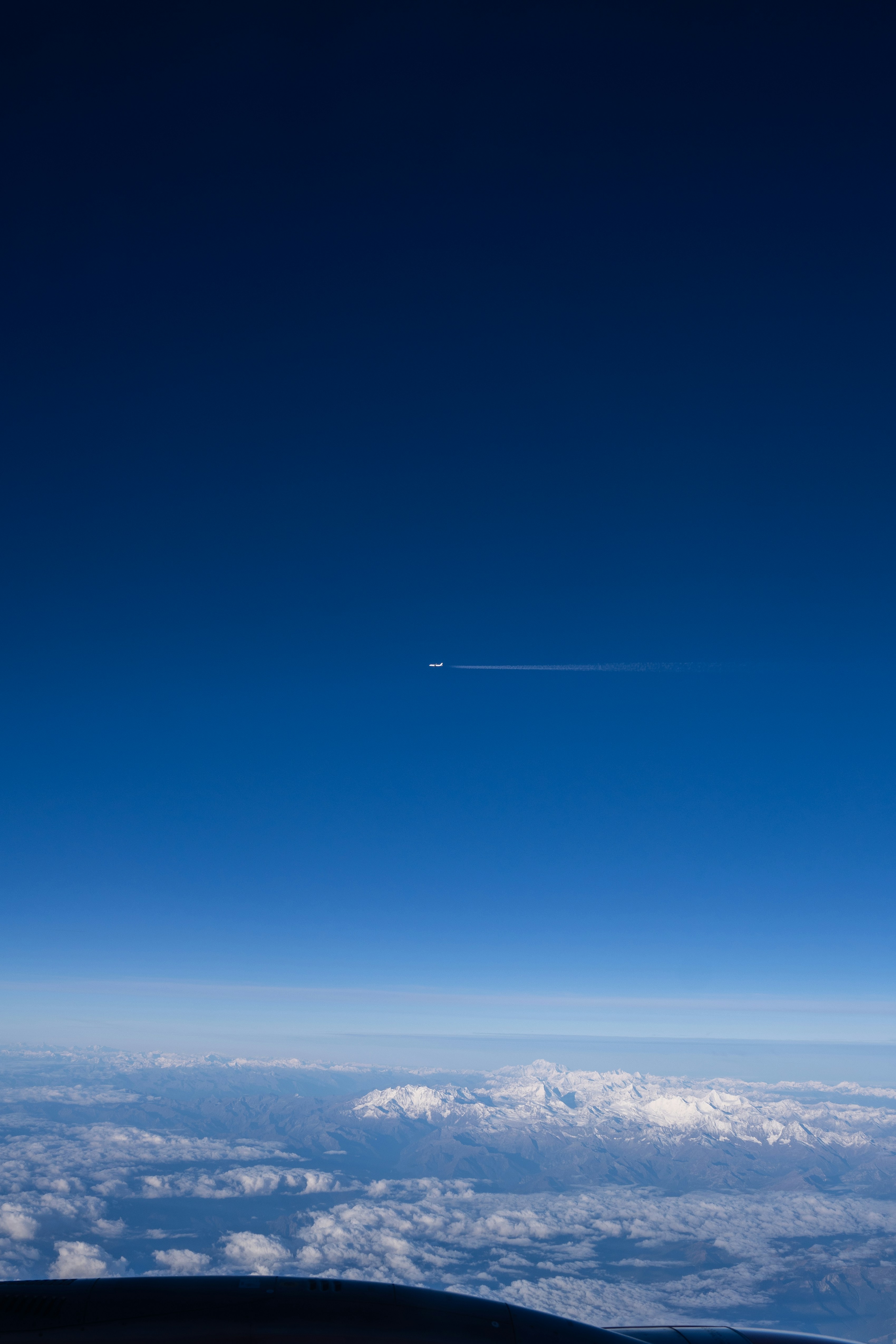A distant airplane leaves a vapor trail against a backdrop of deep blue sky and snow-capped mountains below. The scene captures the vastness of flight.