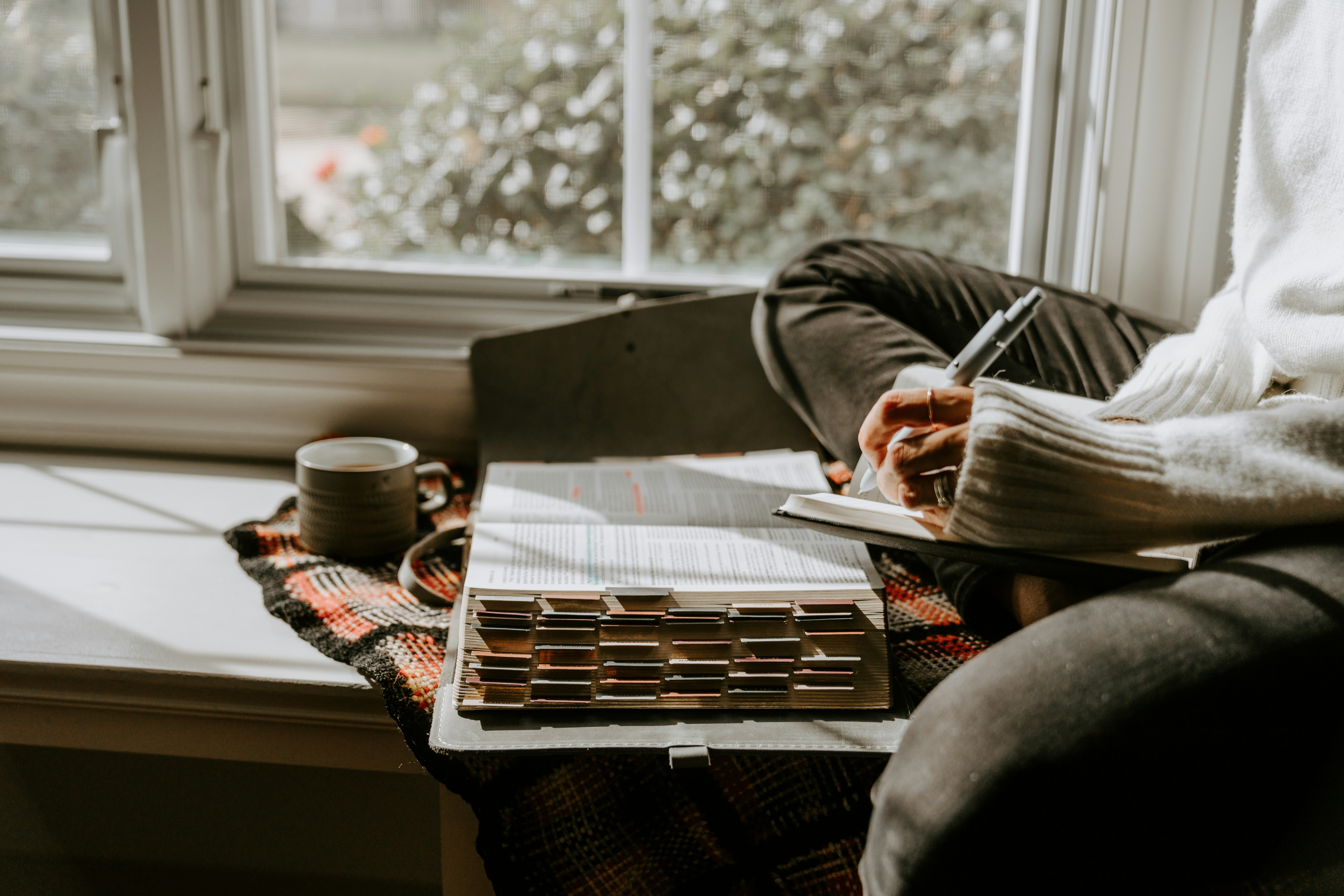 Person writing in a journal with watercolors nearby, seated by a window with soft natural light.