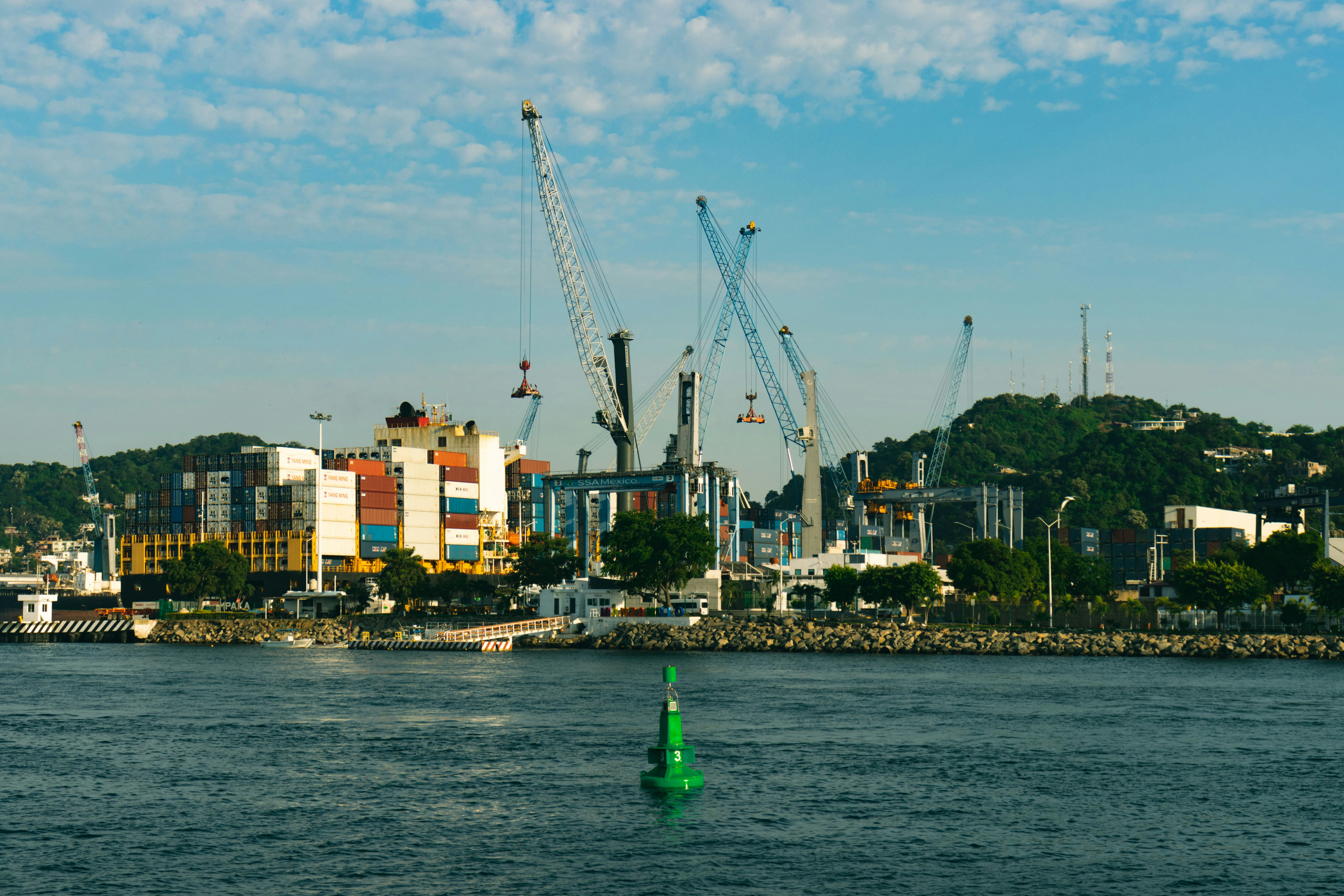 Cranes tower over a bustling waterfront city under a partly cloudy sky.
