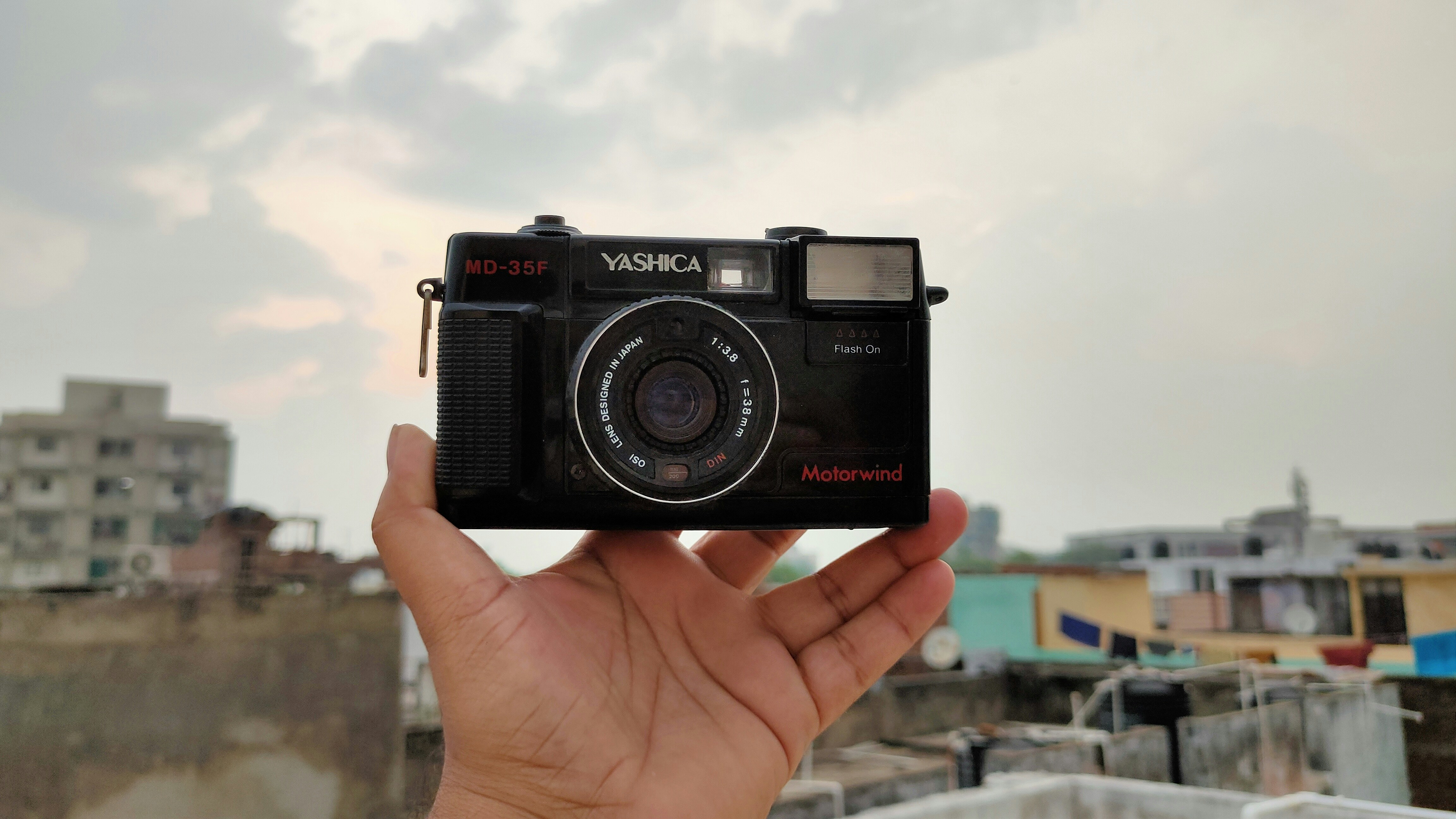 Photograph of a hand holding a vintage Yashica camera with a blurred cityscape in the background.