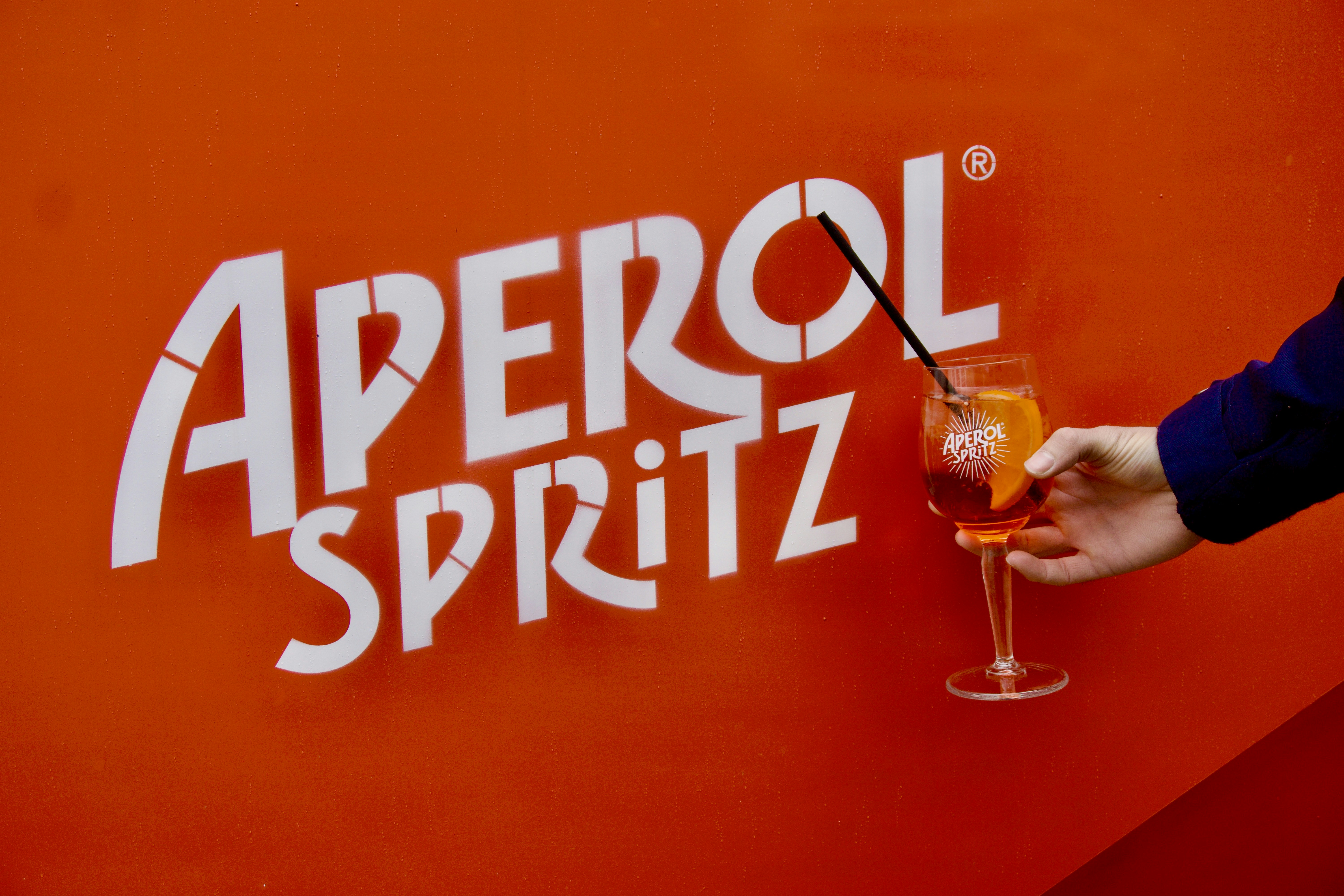 Crimson Wall Toast: A Hand Grasping Aperol Spritz Beside the Logo