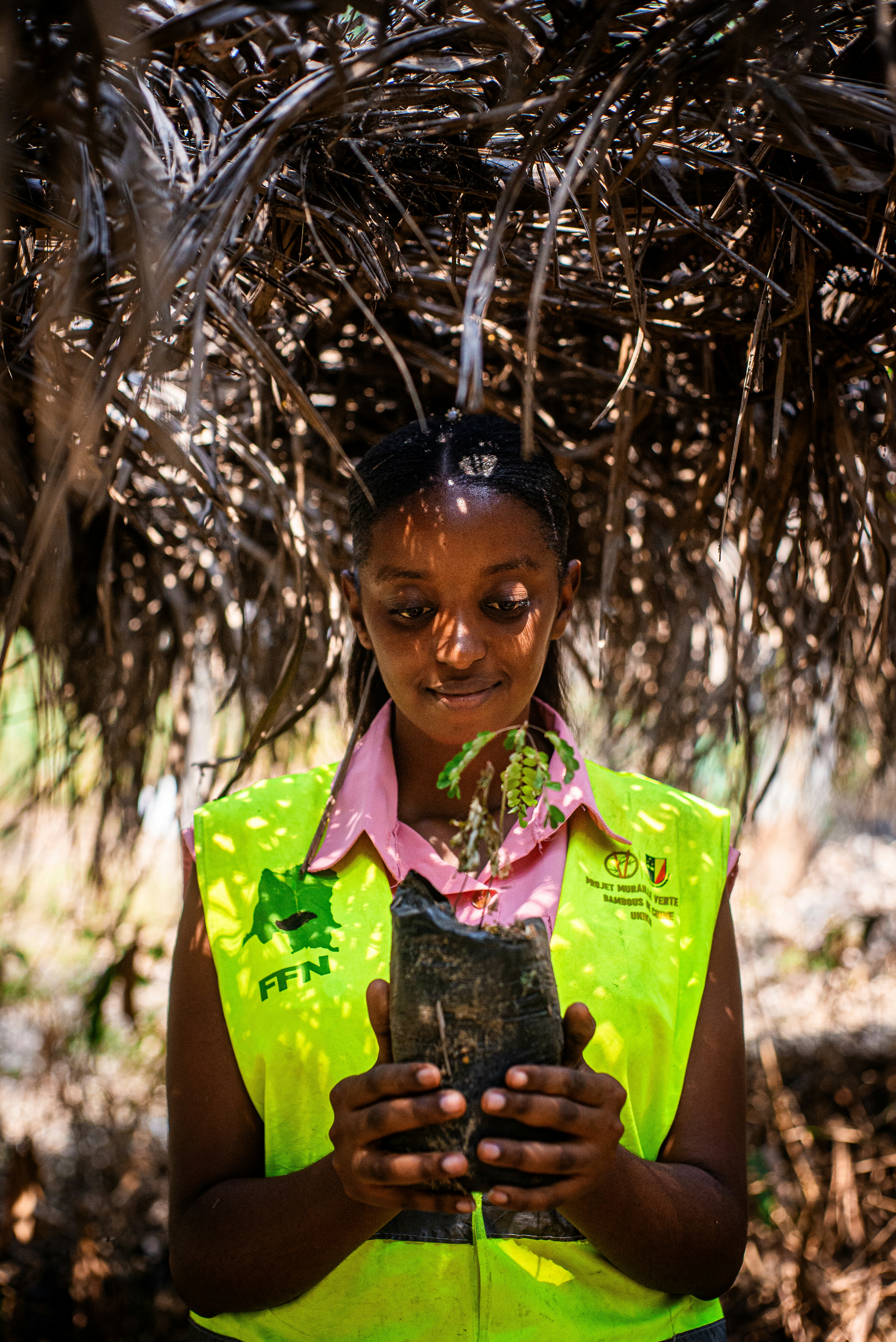 Young woman in a bright vest holds a small plant in a pot, standing beneath a thatched canopy. The scene emphasizes the importance of environmental stewardship.