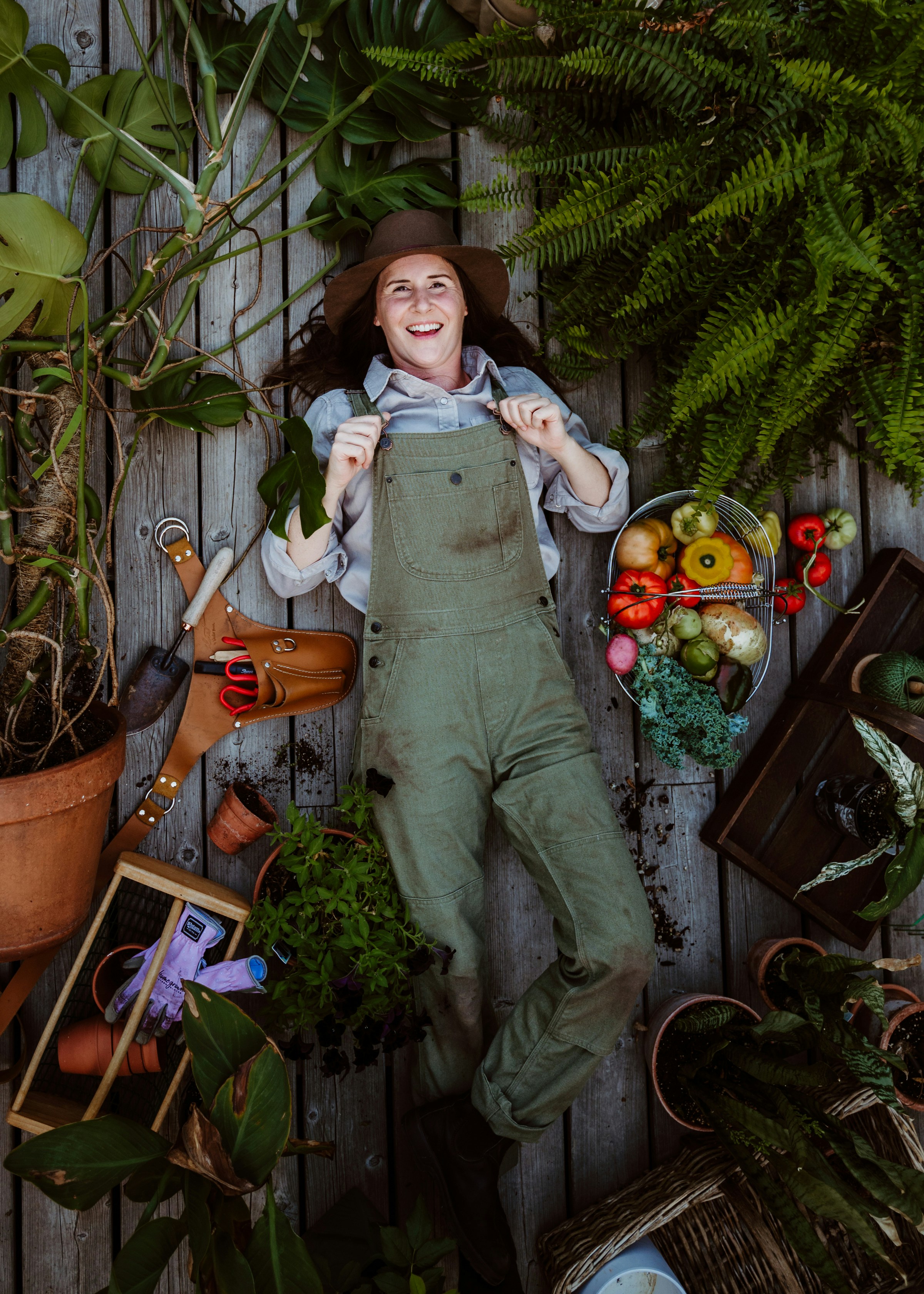 Gardener amongst her tools and plants on the deck smiling wearing green overalls and enjoying being outside working in her garden and with her plants