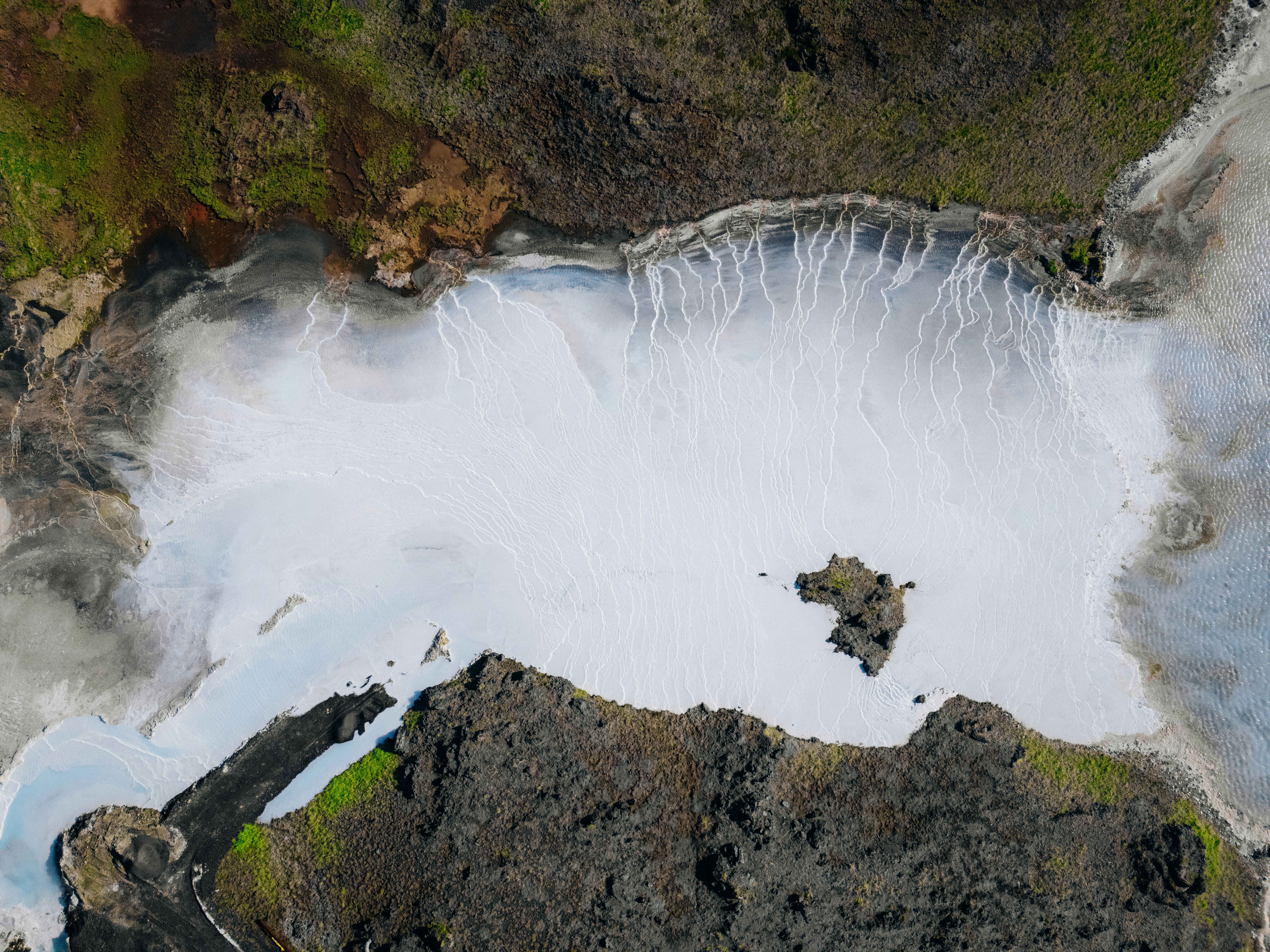 Aerial view of a white salt formation surrounded by rugged terrain, with hints of green vegetation.