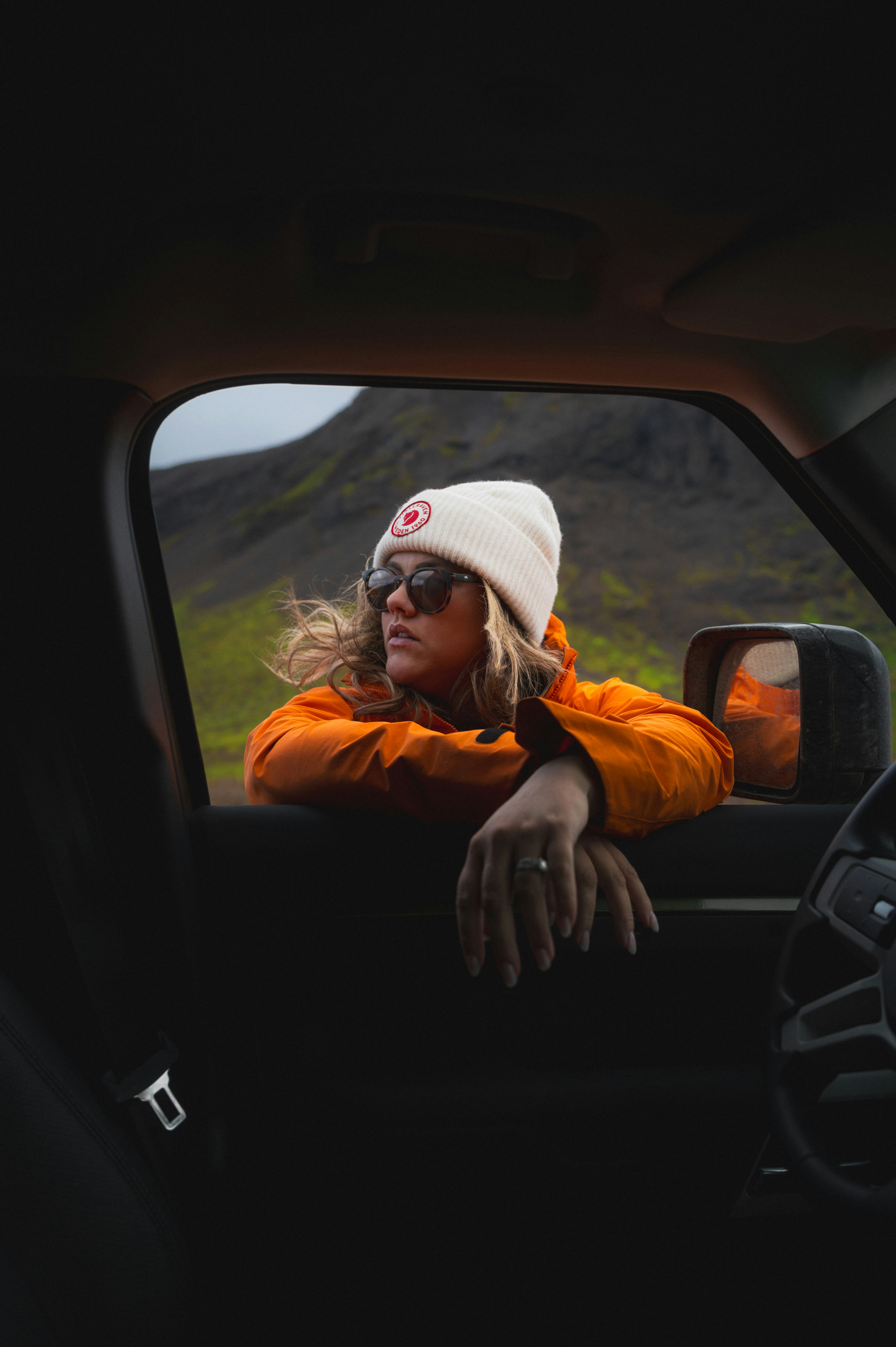 Person in an orange jacket and white beanie leans out of a car window against a rugged landscape.