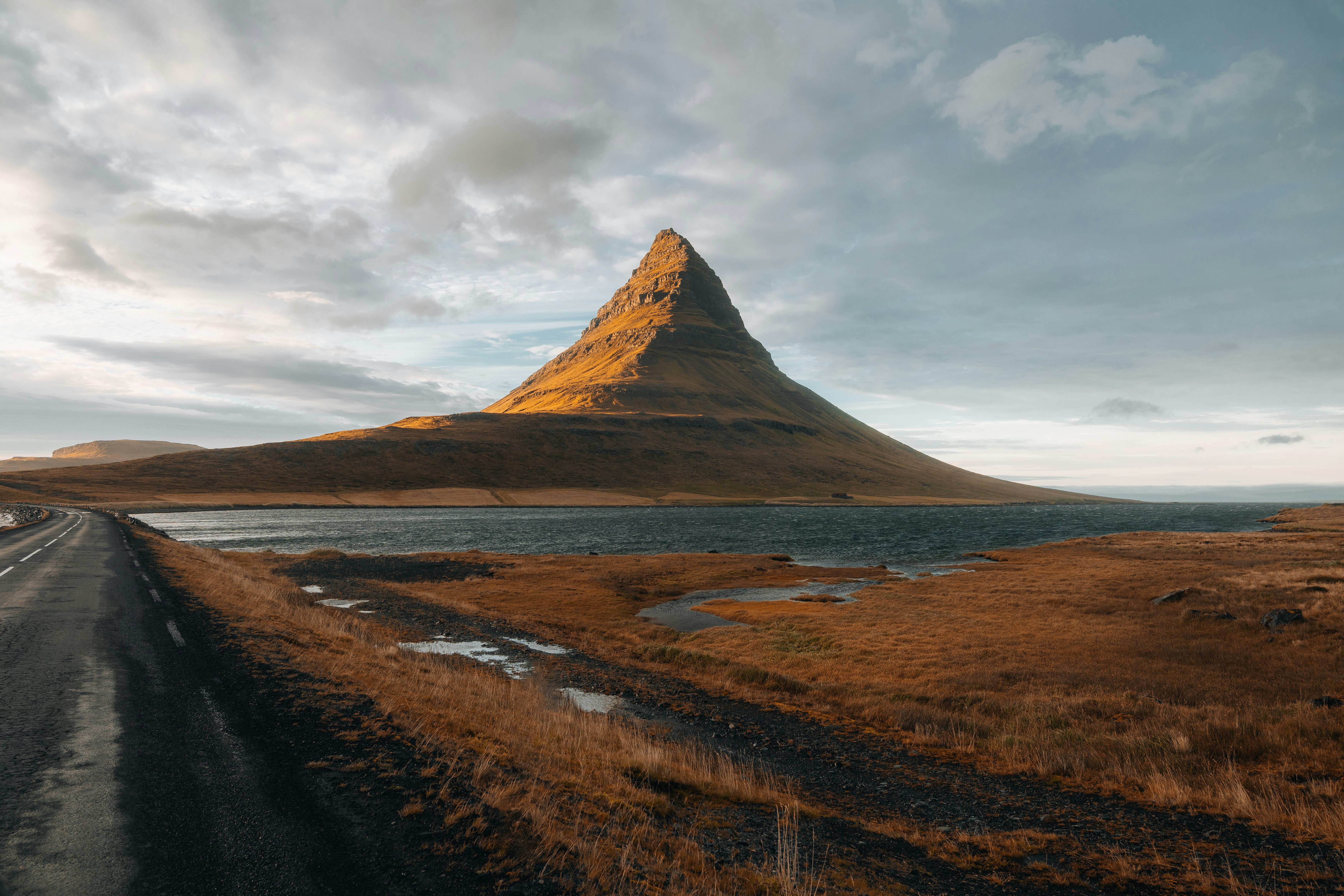 Sunlit Kirkjufell mountain rises sharply beside a winding road under a vast sky.