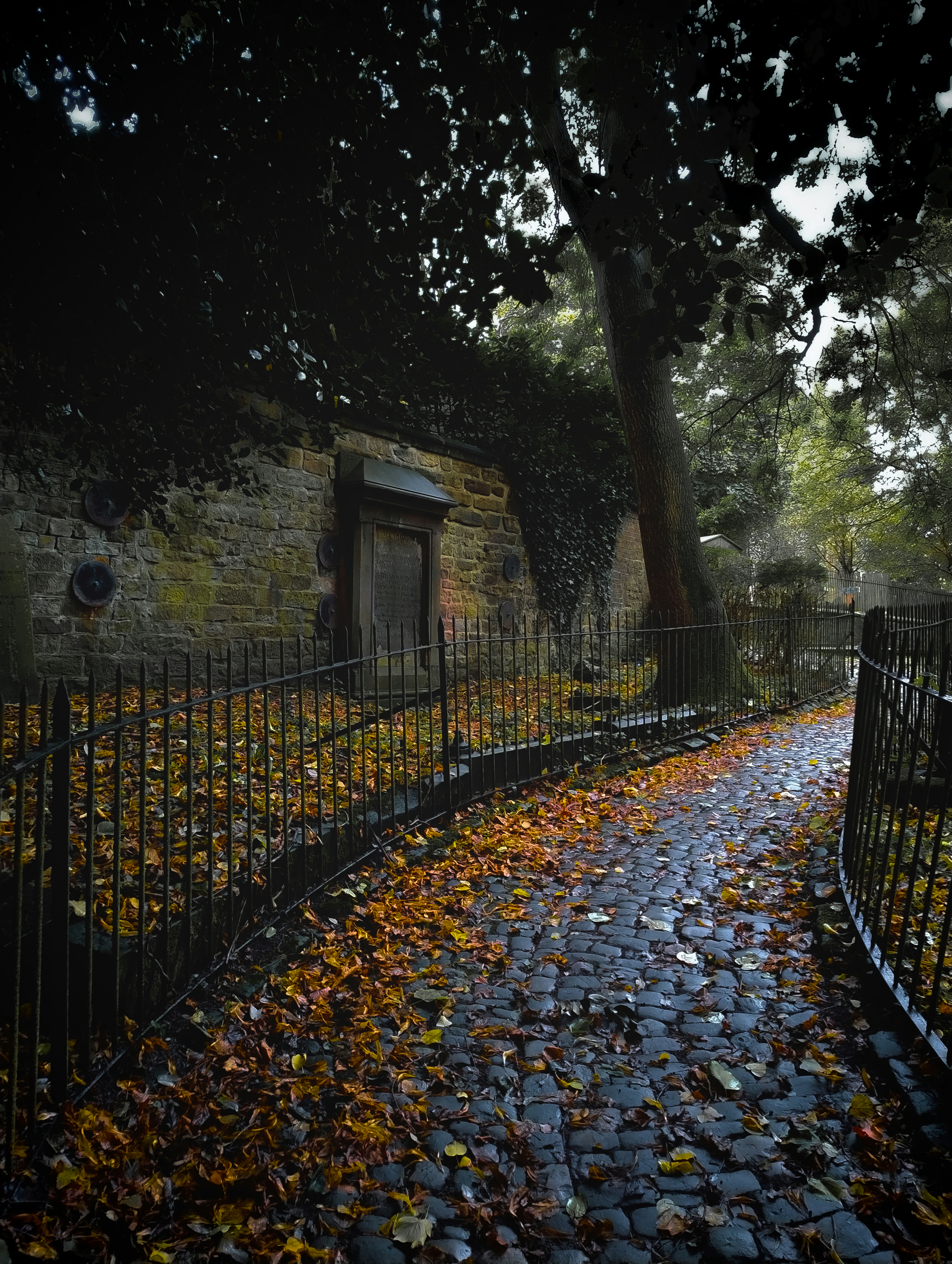 A rain-slick, leaf-strewn cobblestone path curves between wrought-iron fences toward a weathered doorway set in a mossy stone wall, framed by dark trees.