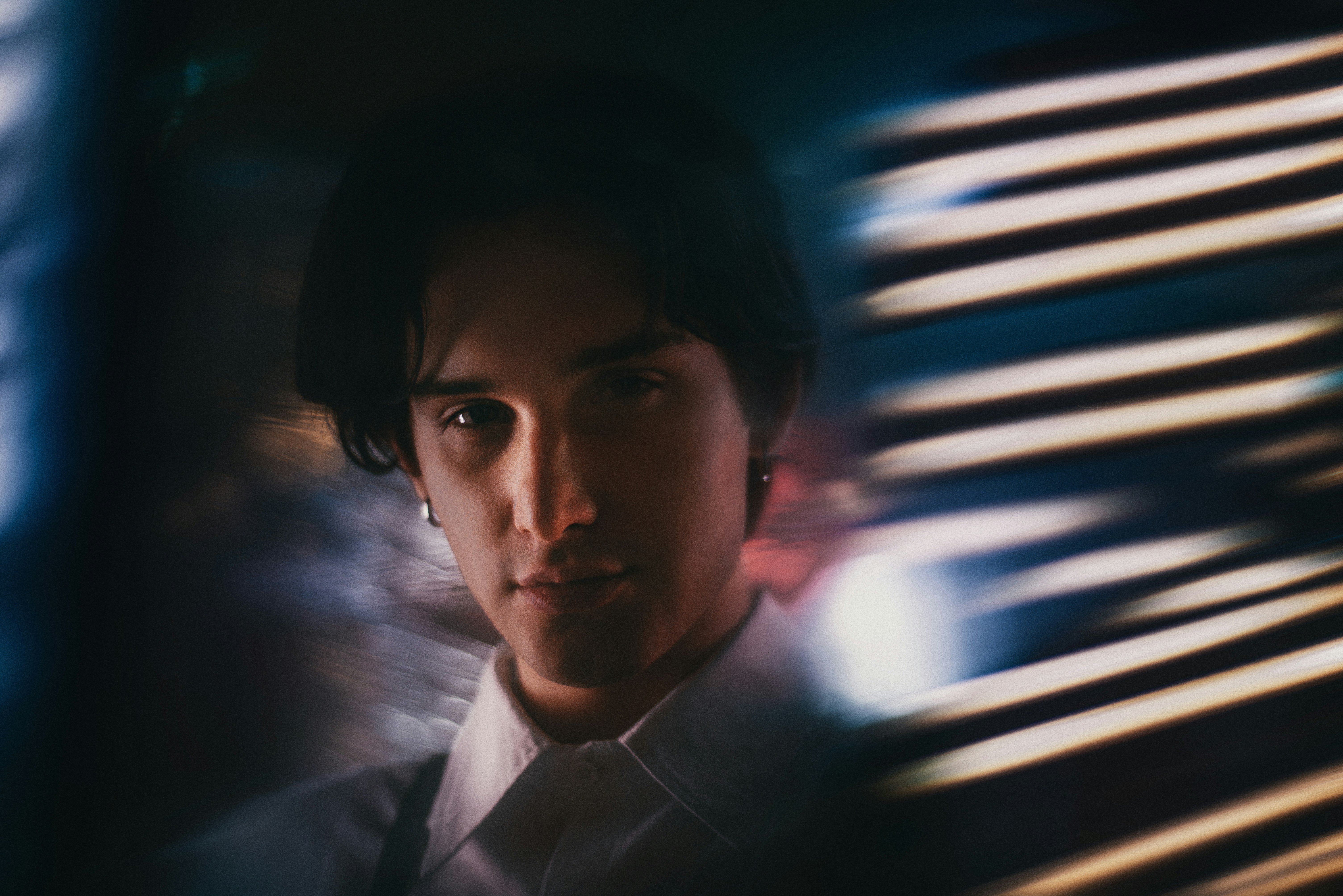 Portrait photograph in soft, low light with diagonal light stripes across the frame, highlighting a quiet gaze.