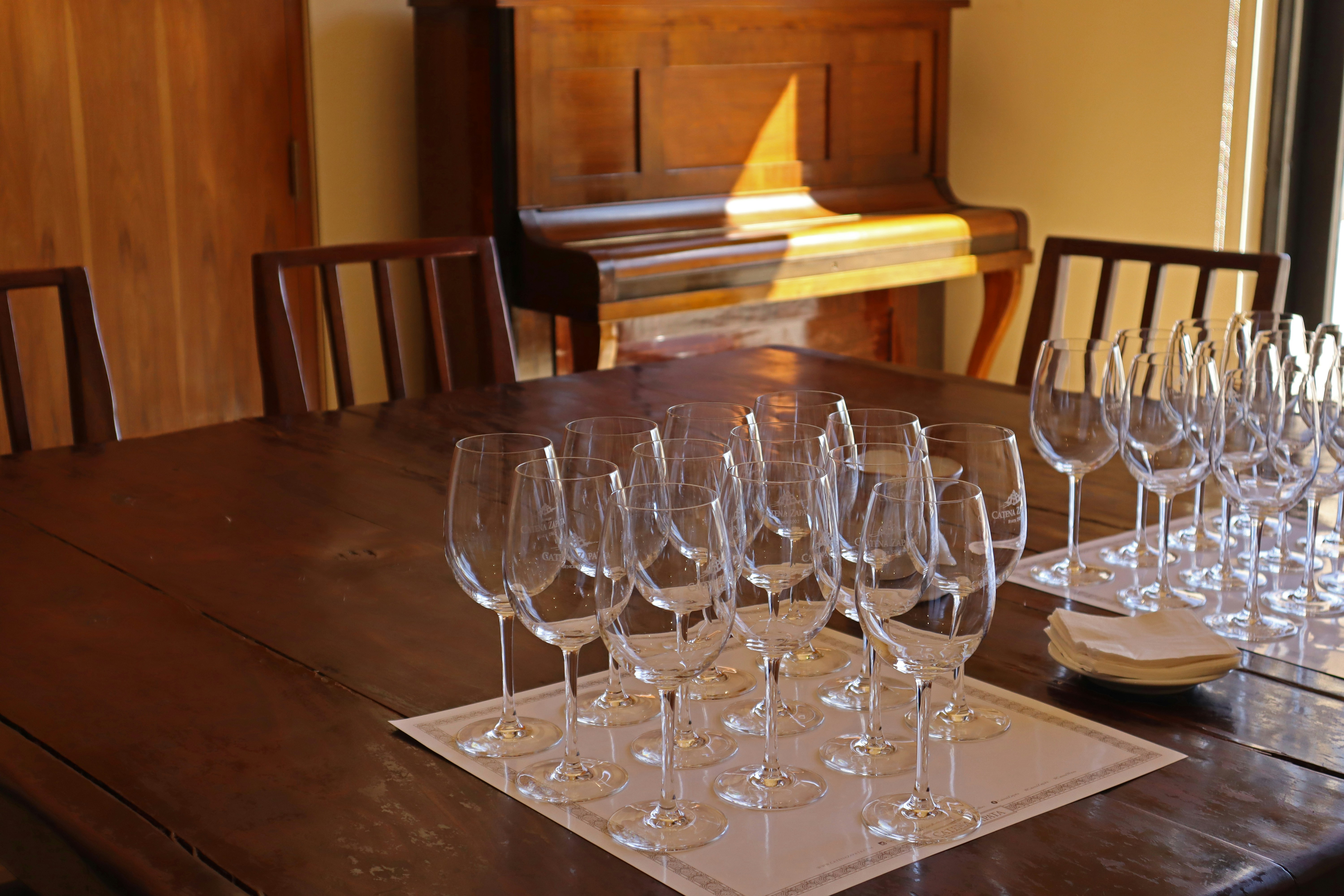 Array of wine glasses on a wooden dining table with a piano in the background, bathed in warm sunlight.