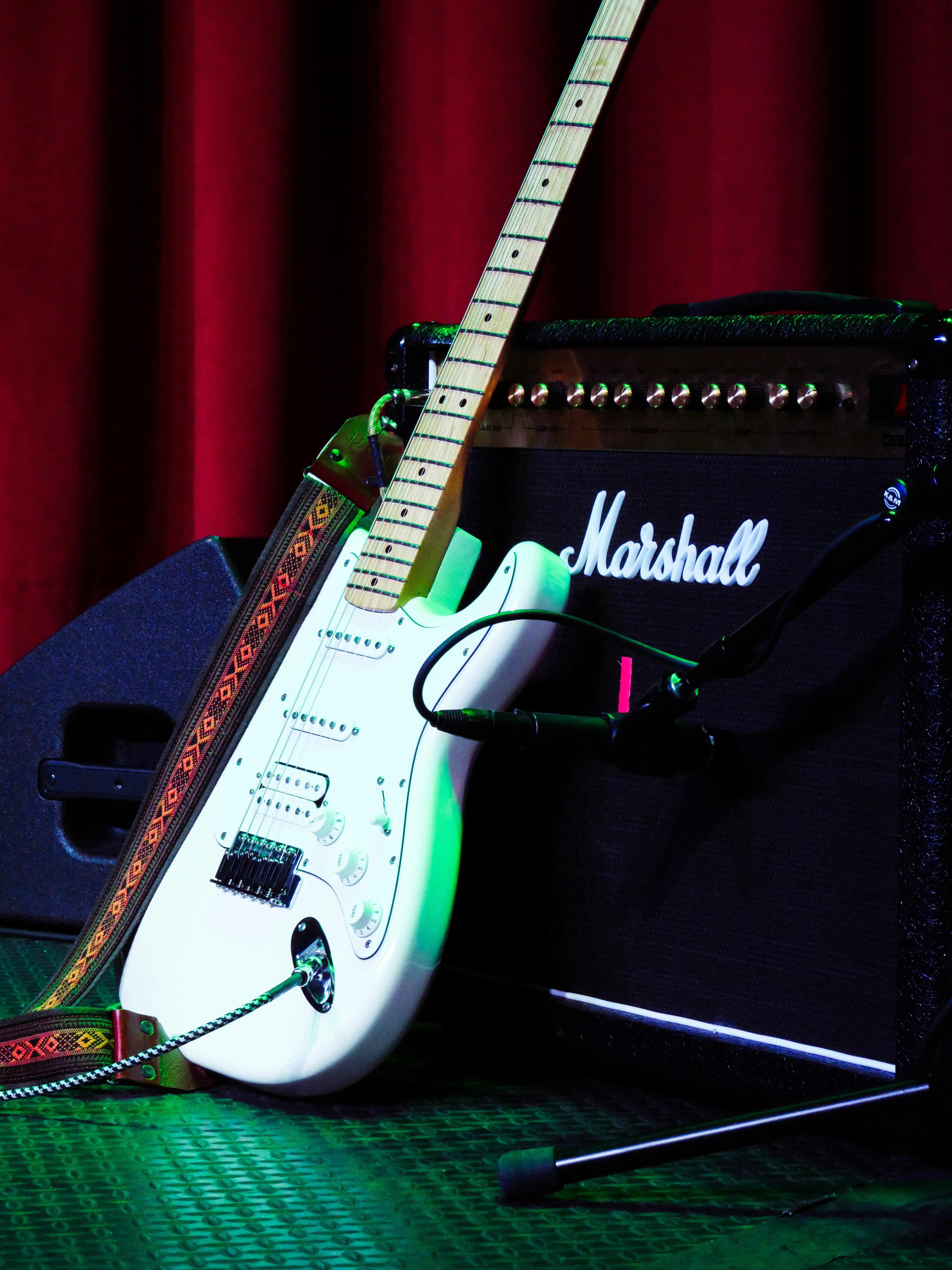 Photograph of a white Tele-style electric guitar leaning diagonally against a Marshall amplifier on a stage with red curtains, bathed in neon-green light.