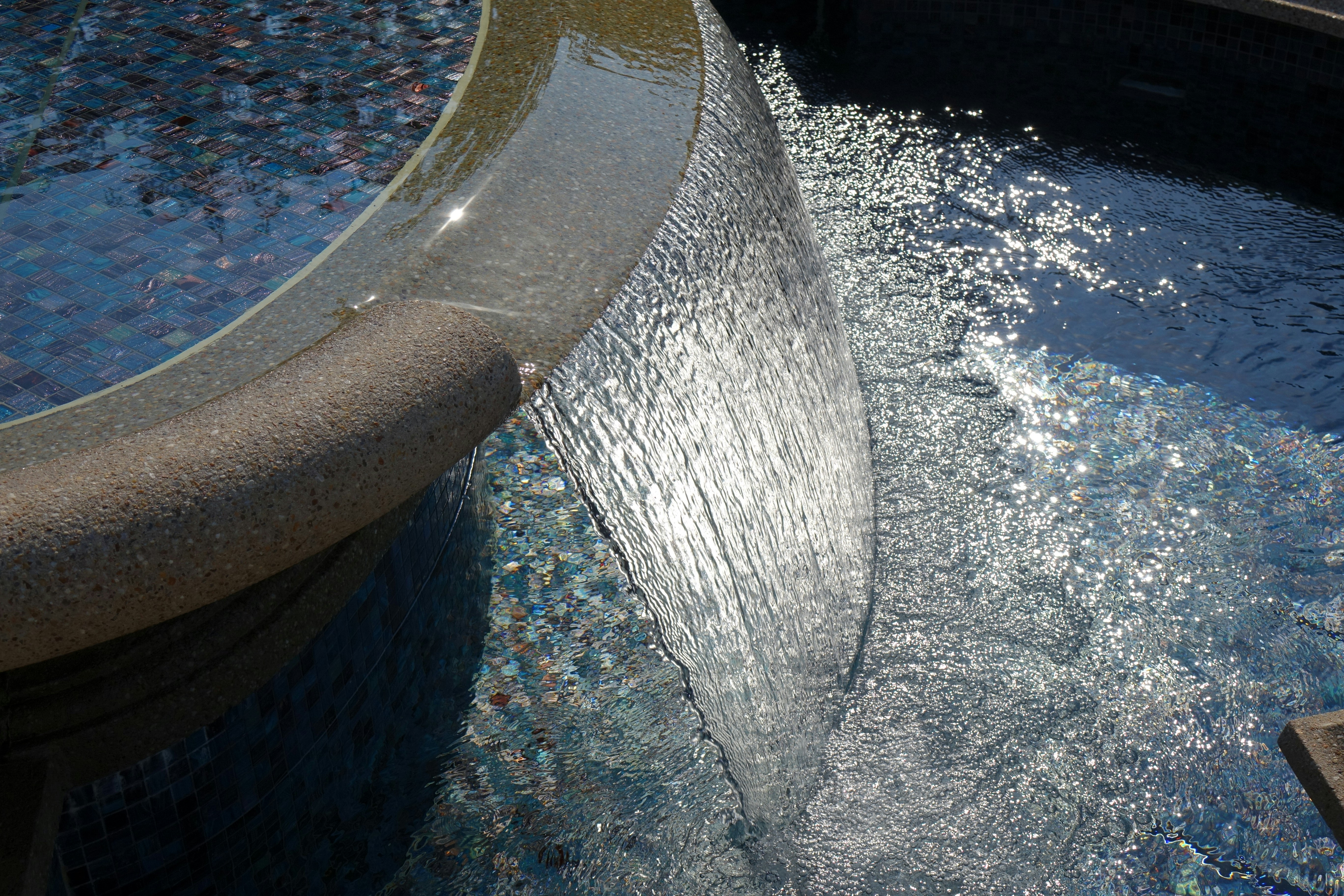 Curved stone spillway releases water into a blue tiled pool, creating a shimmering curtain and rippled surface.