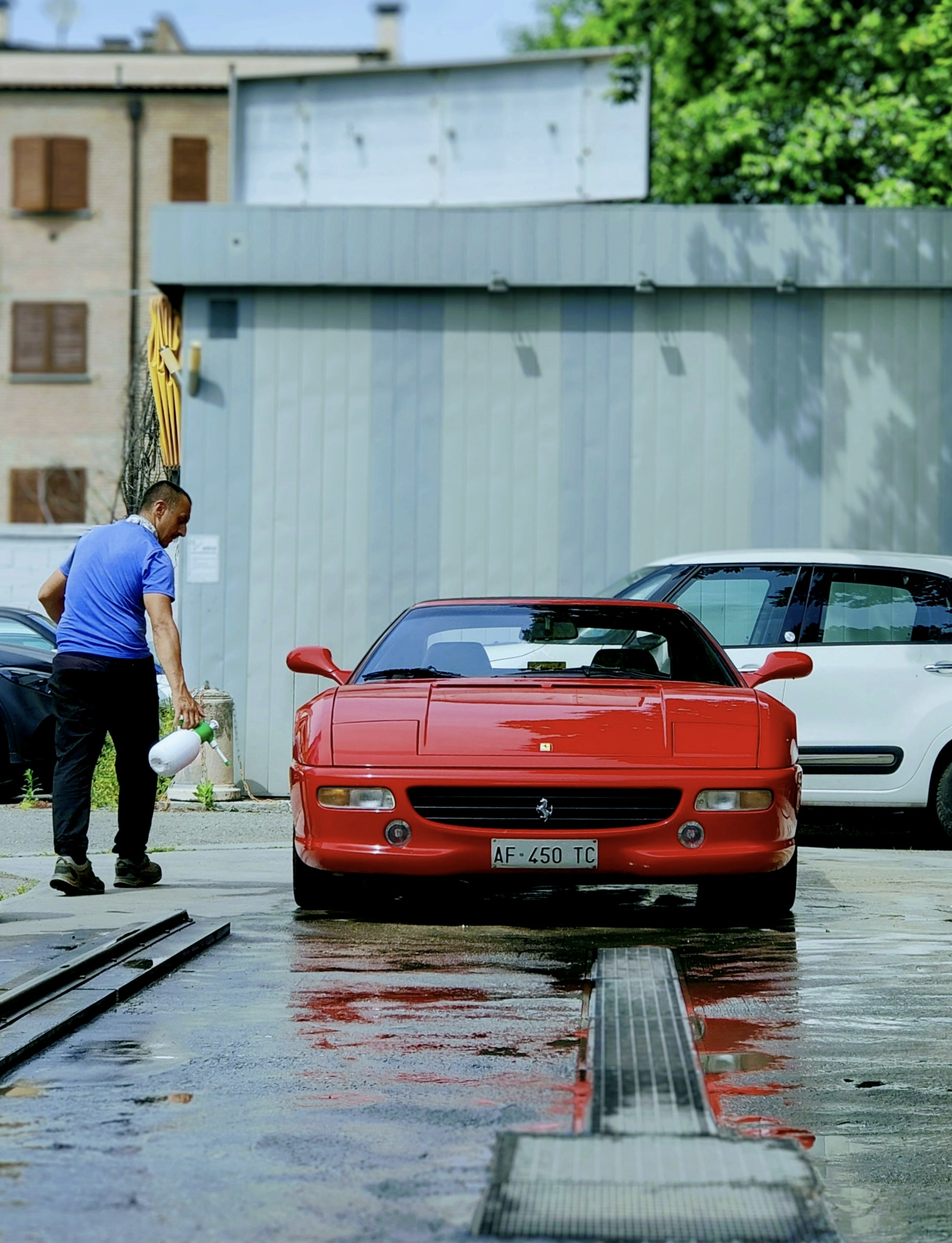A red Ferrari convertible is being washed by a man, with wet pavement reflecting the car against a gray industrial backdrop.