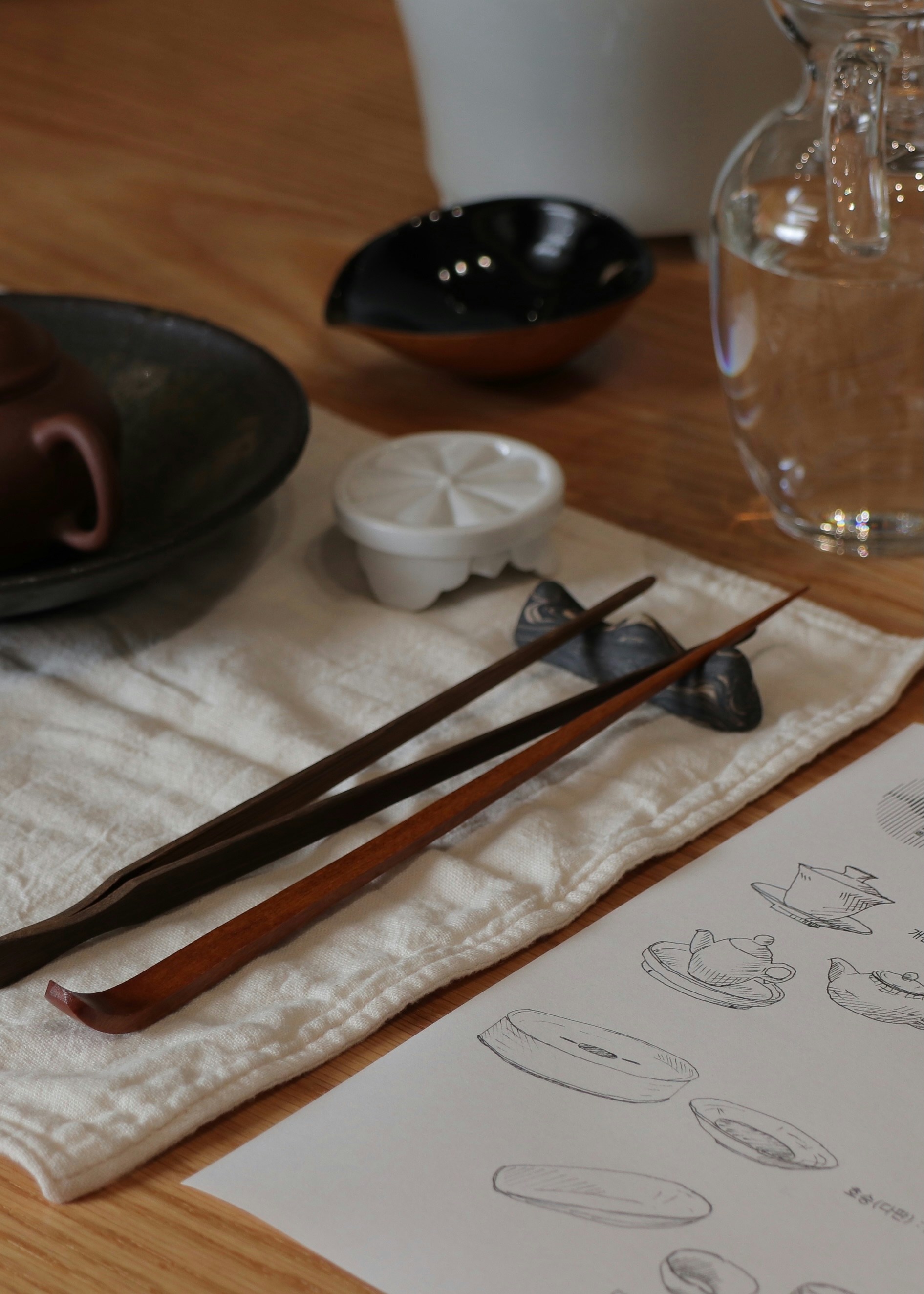 A still-life photograph featuring lacquered chopsticks resting on a white napkin, with ceramic vessels and a sketch sheet on a wooden table.